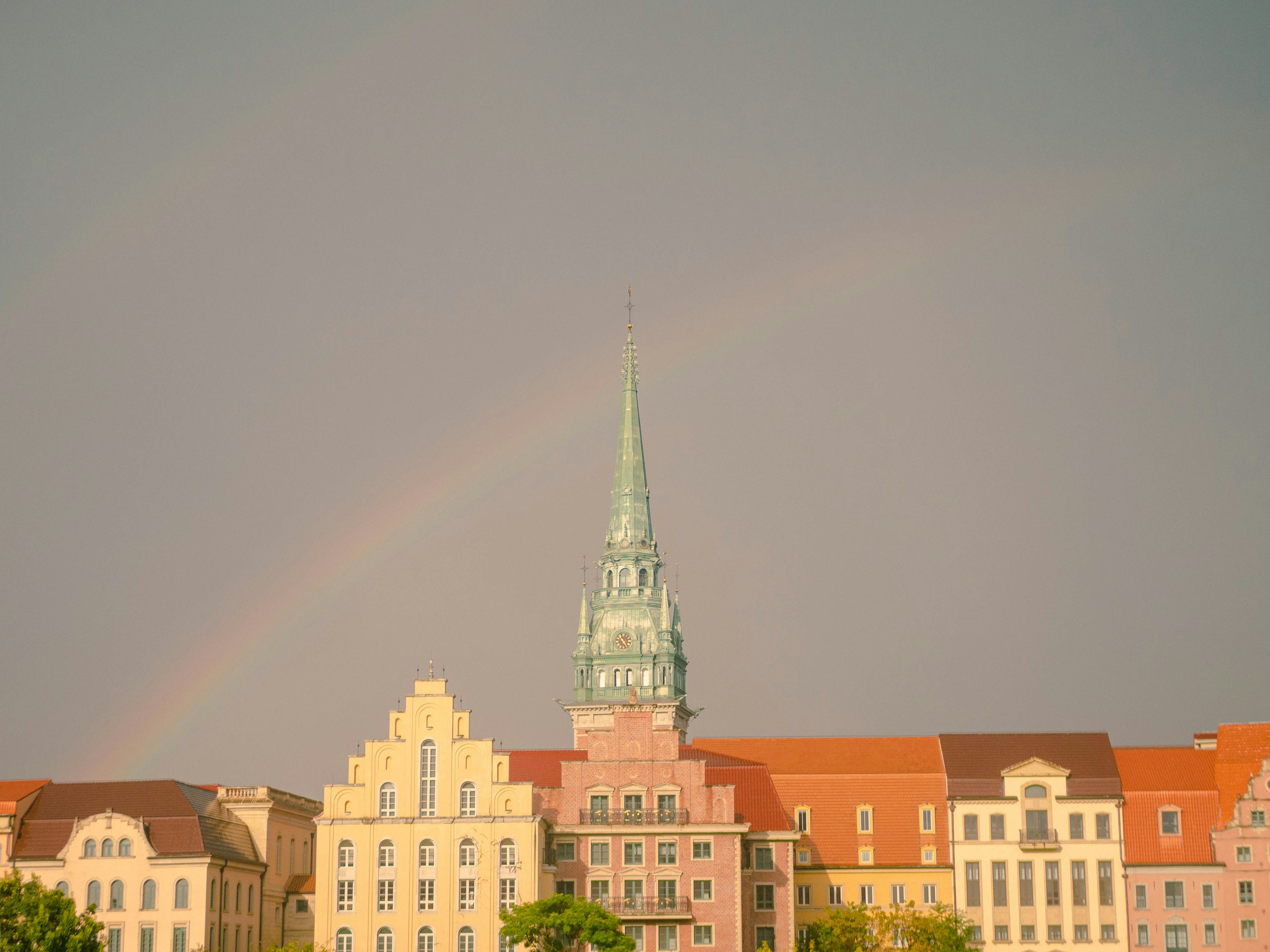 Pastel-colored city rooftops rise beneath a soft rainbow, with a tall green-spired church dominating the skyline.
