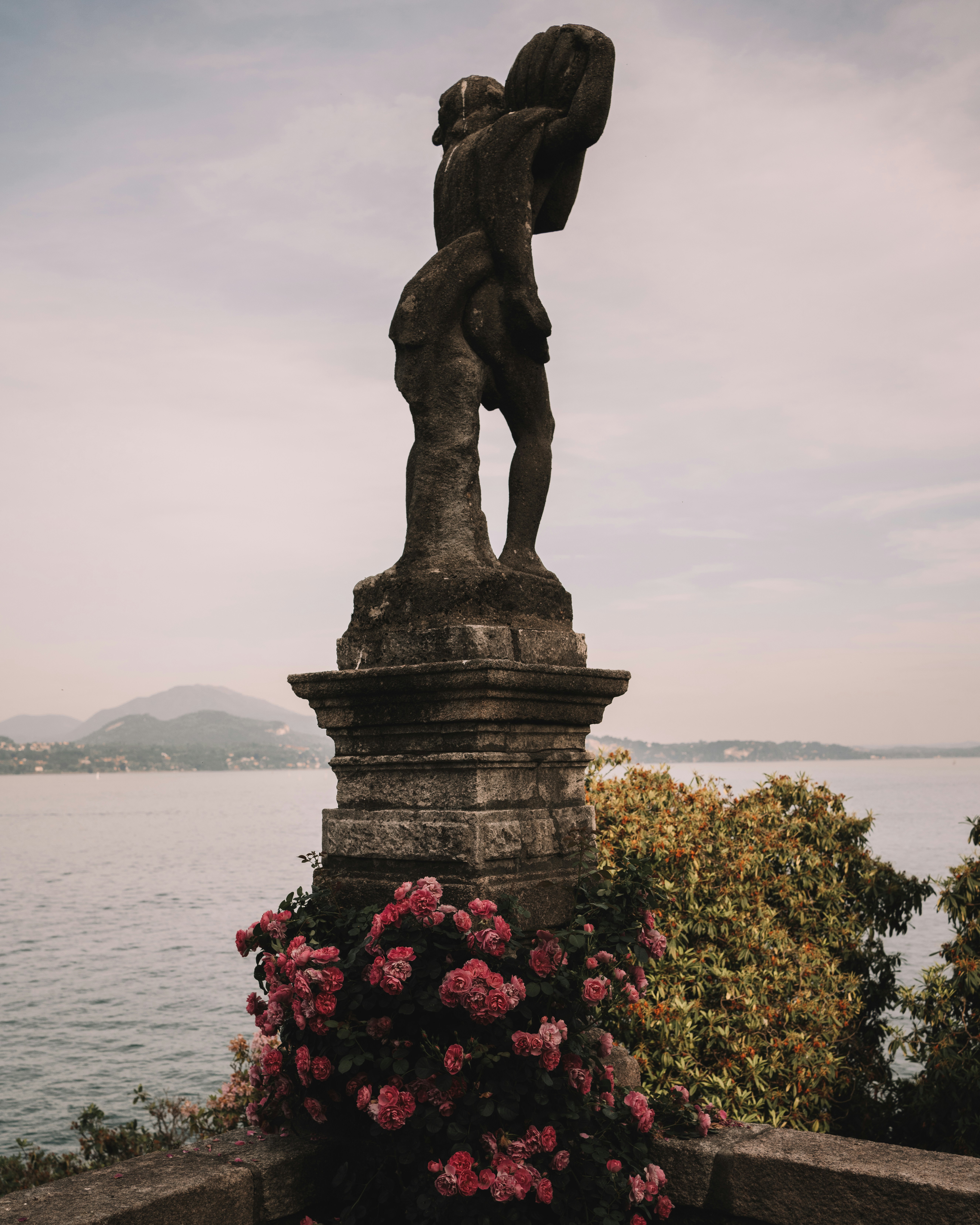 Floral Sentinel by the Lake: A Weathered Statue Amid Pink Blooms