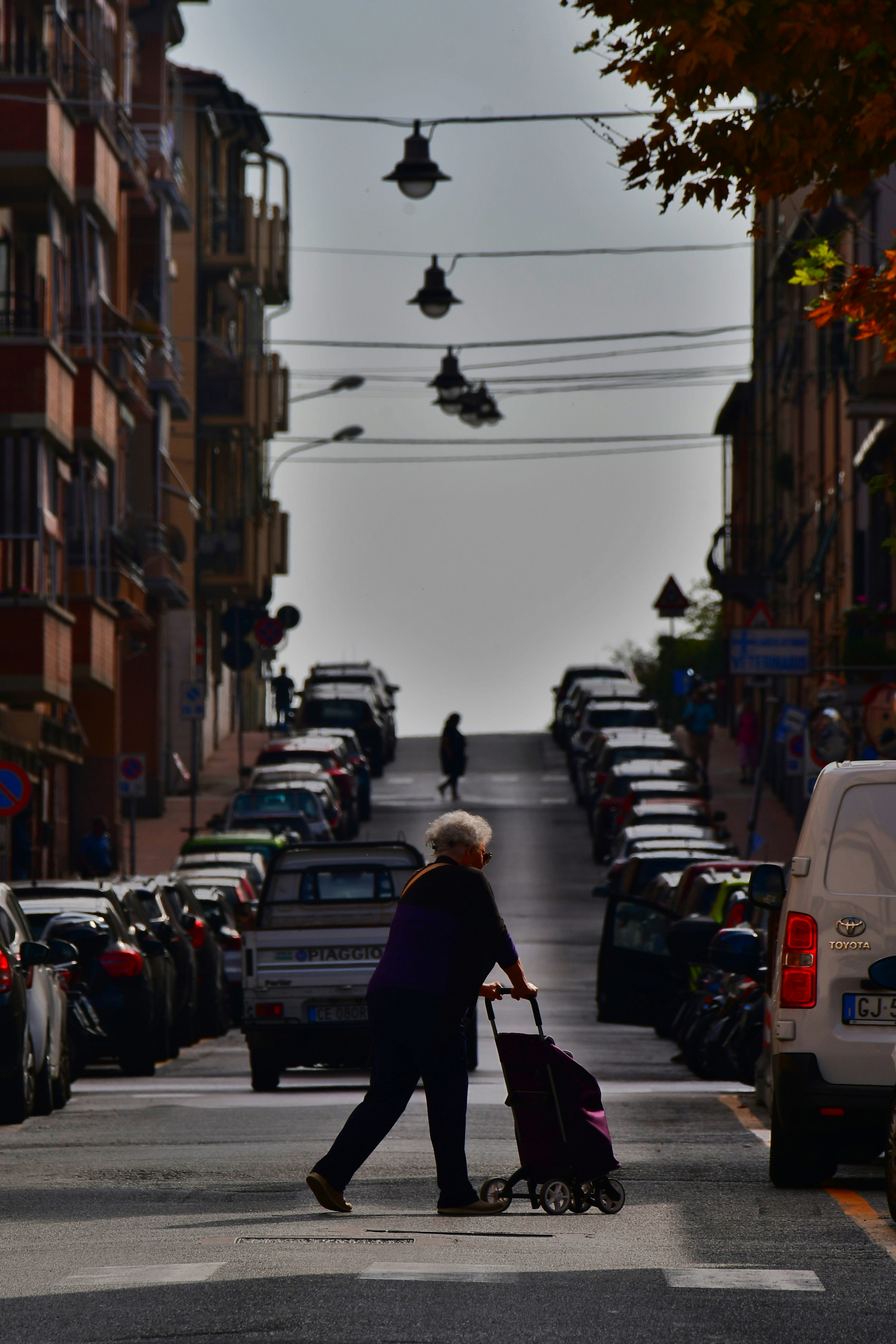 Elderly person crossing a steep, car-lined street with overhead streetlights.