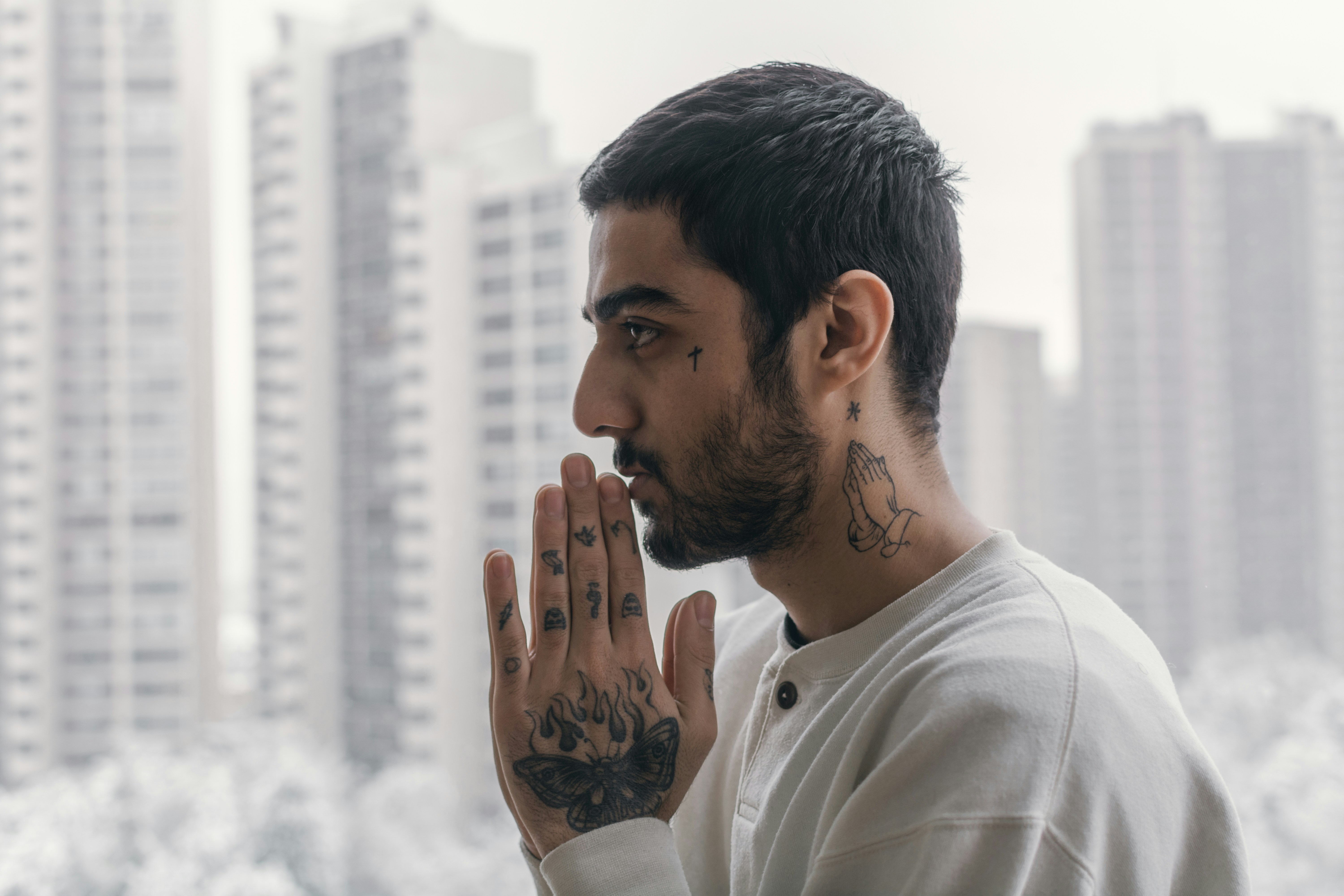 A thoughtful portrait of a tattooed man gazing out at a cityscape, offering a striking contrast between the intricate tattoos and the blurred high-rise buildings in the background. The muted color palette of grays and whites creates a serene atmosphere, highlighting the subject's introspective mood. This image captures the balance between urban life and personal reflection, making it visually compelling.