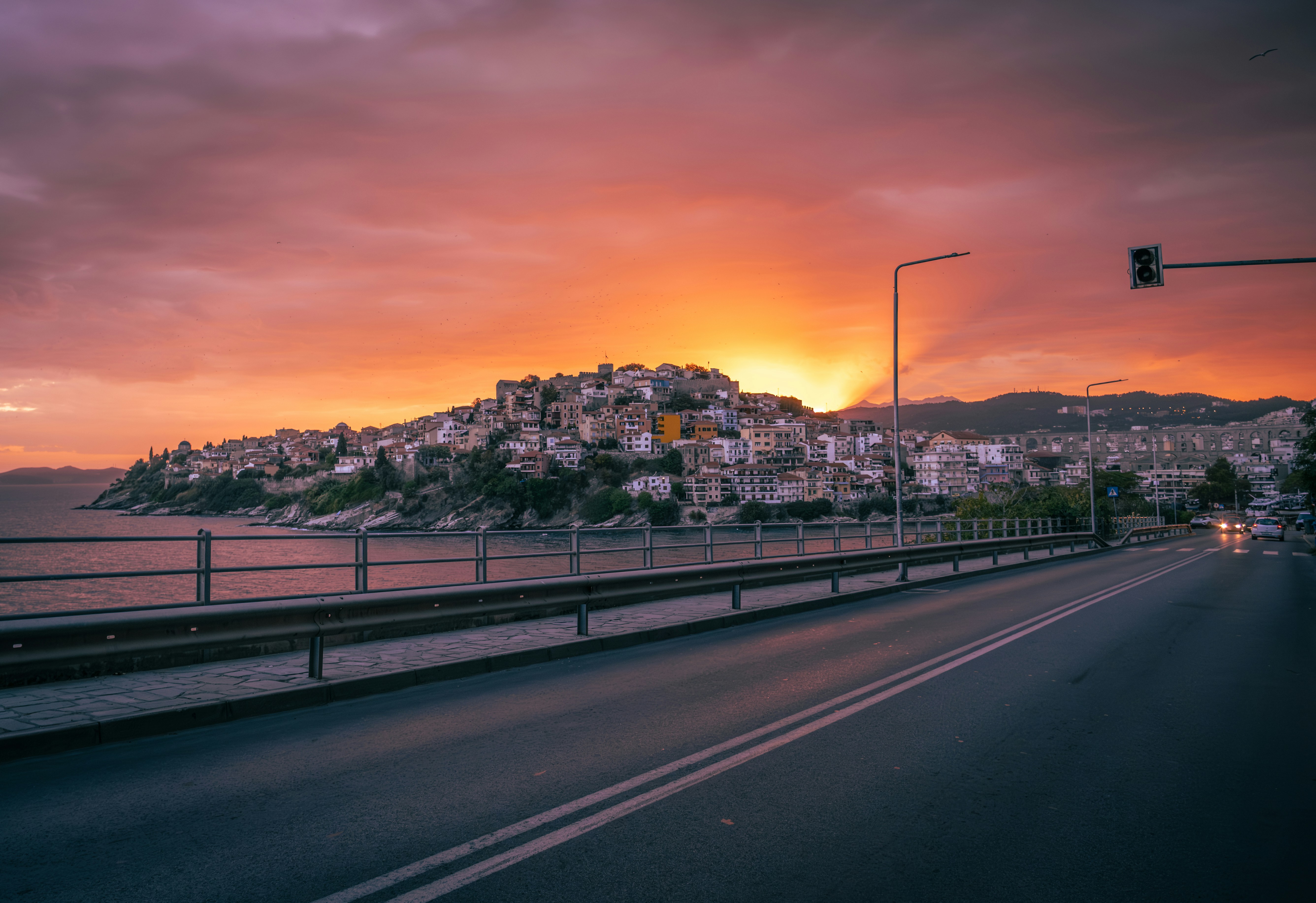 Sunrise illuminates a hillside town beside a calm sea, with vibrant orange and purple hues in the sky.