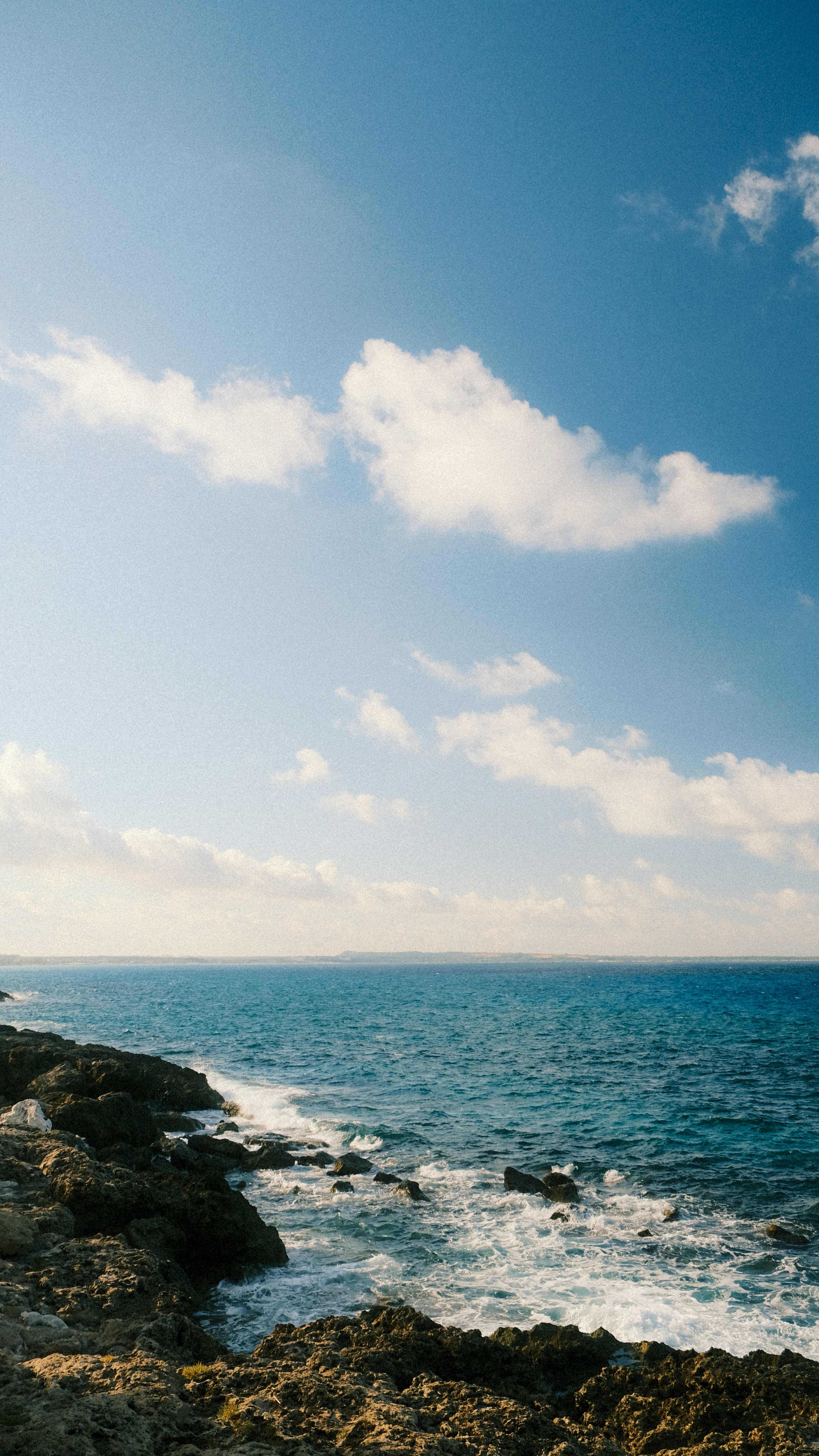 Person seated on a rocky shoreline gazing at the expansive ocean under a bright blue sky.
