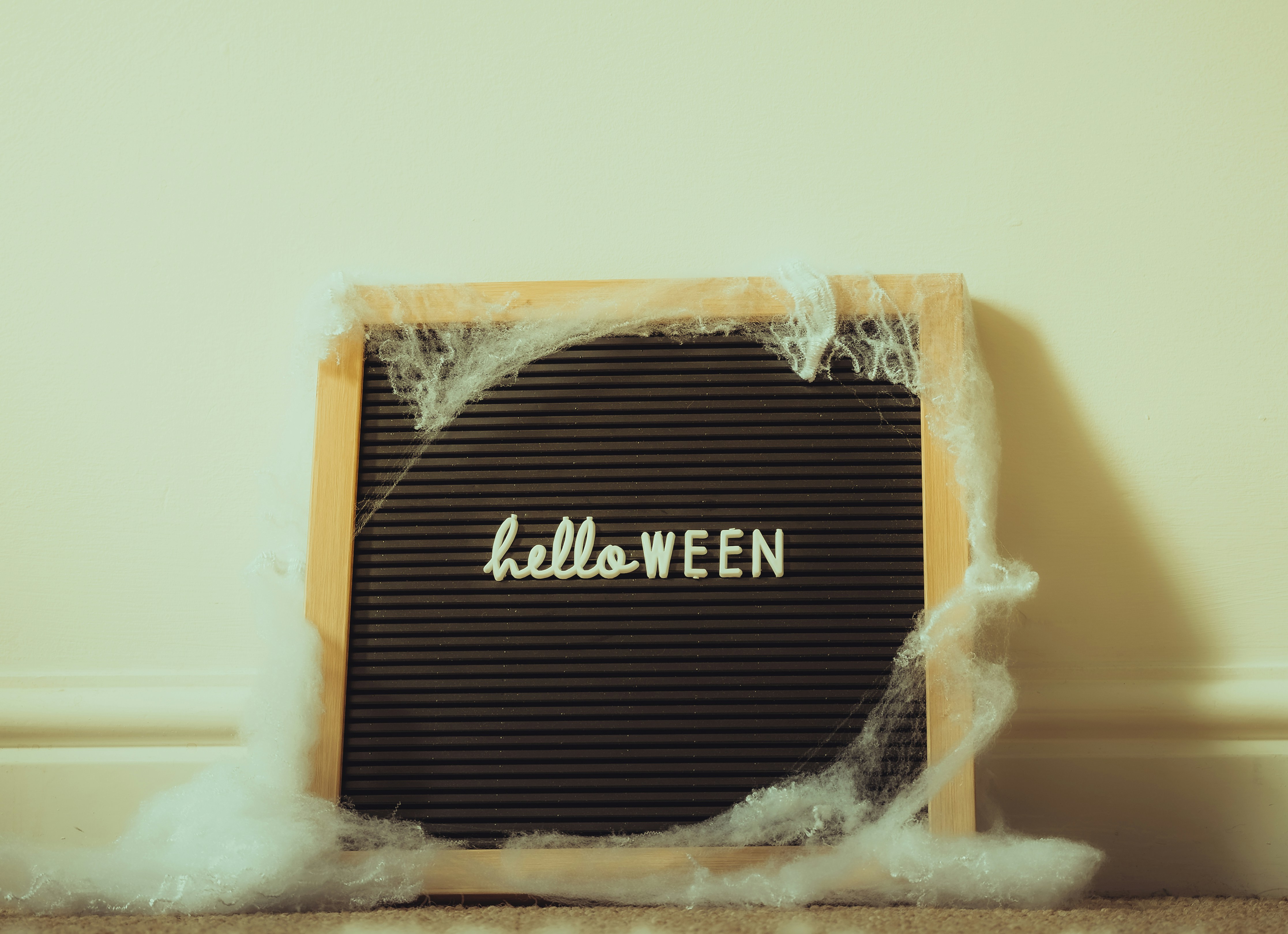 A small slate sign reading 'helloWEEN' sits centered on a wooden frame, surrounded by spider webs along a pale wall and baseboard. An autumnal Halloween display intended for a home decor scene.