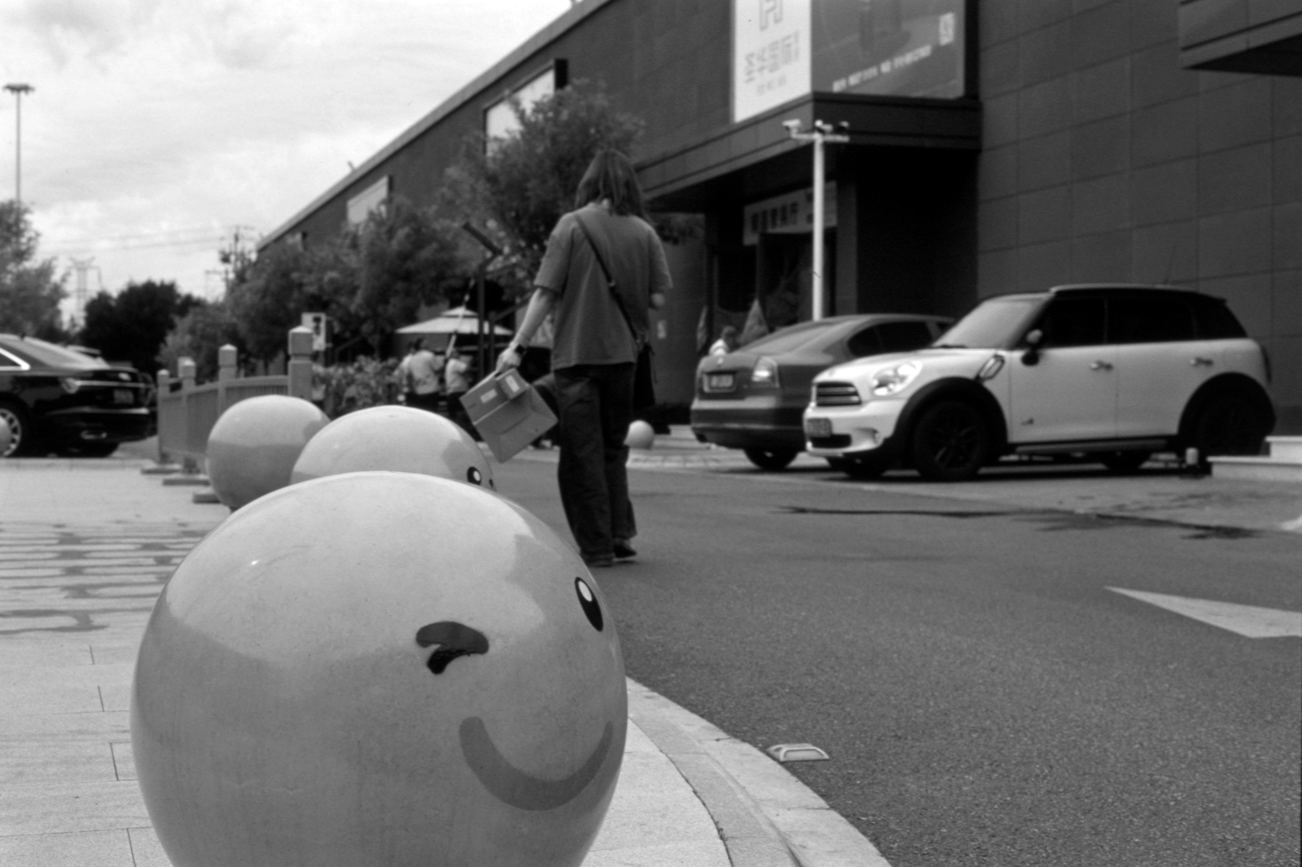 Black and white street scene with a smiling bollard in the foreground and a man walking towards a parked car.