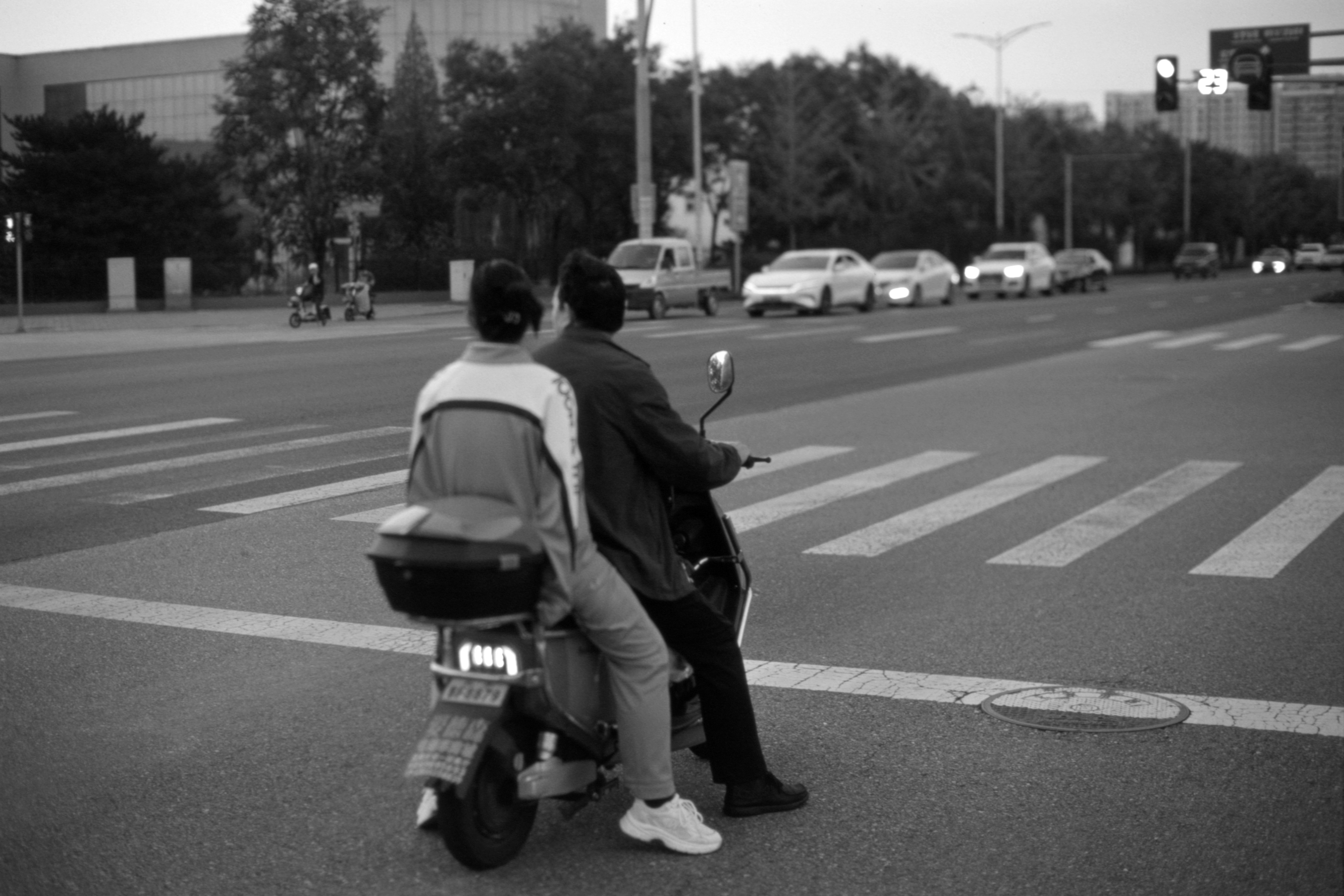 Photograph of two scooter riders at a zebra-crossing in an urban street, captured in black and white.