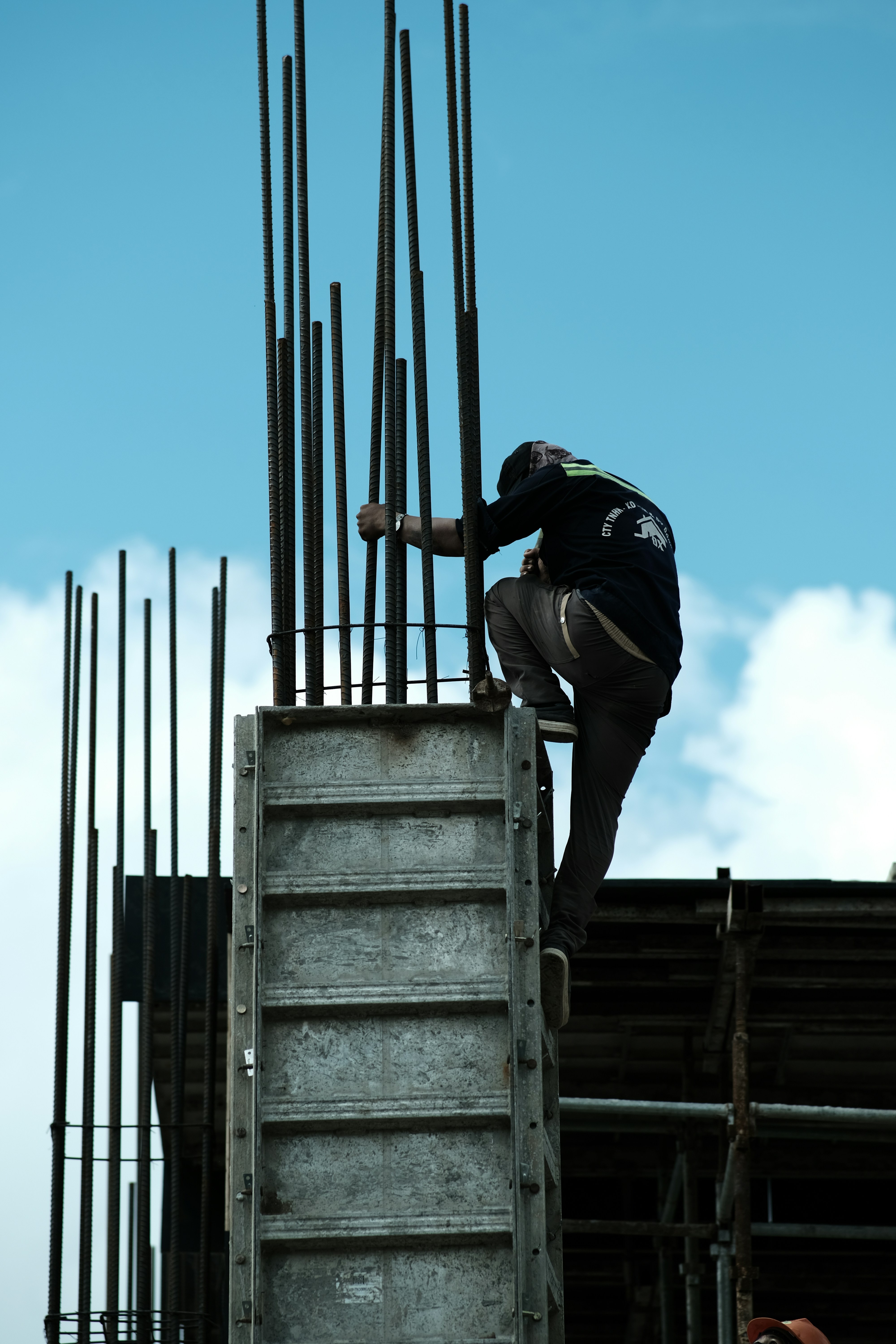 Construction worker climbs a concrete formwork framed by vertical steel rebar against a bright blue sky.