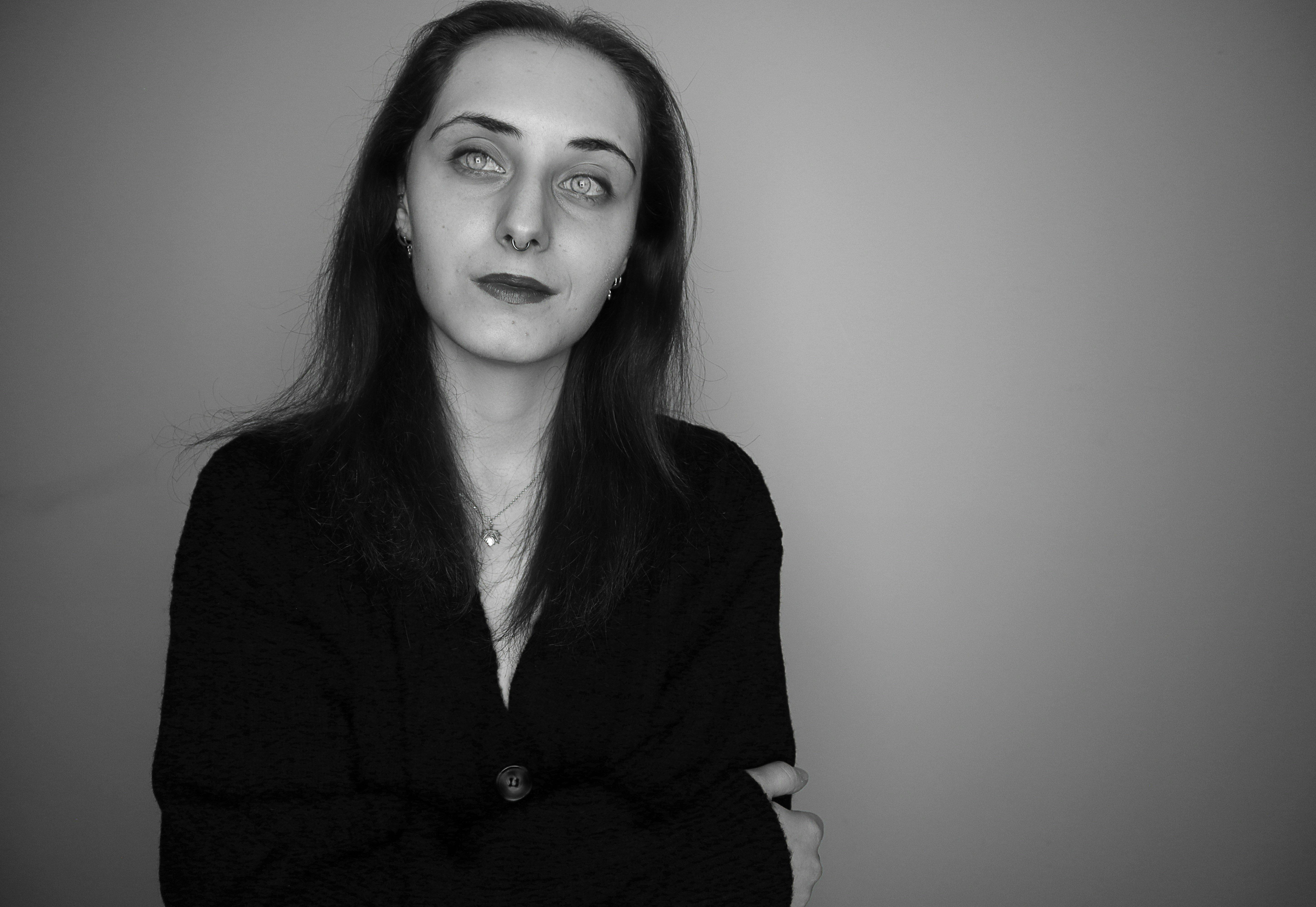 This striking black and white portrait captures a serene expression, highlighting the subject’s piercing gaze and subtle features. The composition focuses on the upper body against a plain background, drawing attention to the contrast between the subject's dark attire and the soft, even lighting. The grayscale palette enhances the mood of introspection, creating a timeless and evocative image.