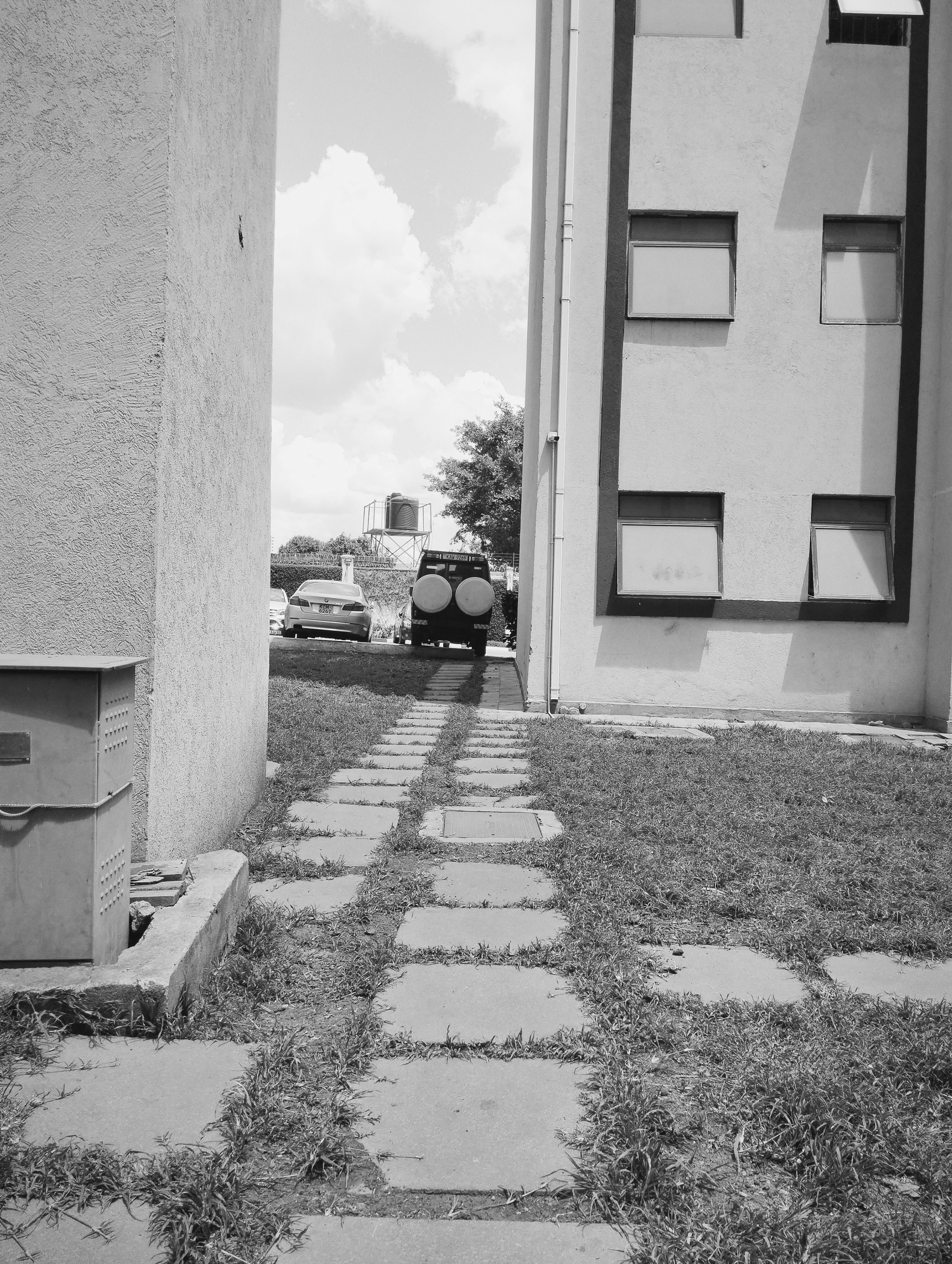 Narrow pathway lined with stone slabs between two buildings, leading towards a parked vehicle under a cloudy sky.