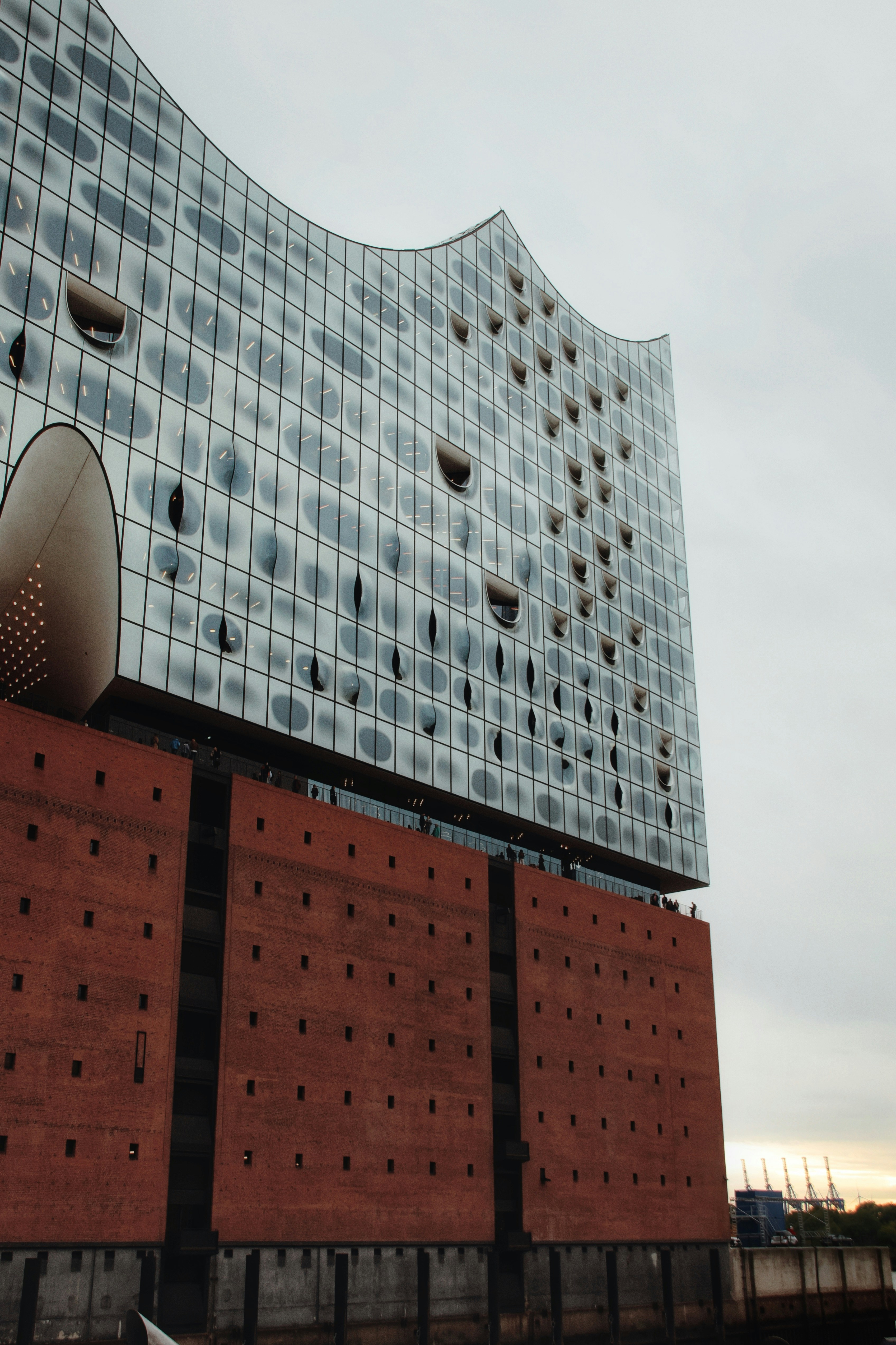 Photograph of the Elbphilharmonie's glass facade curving above a red brick base against a pale sky.