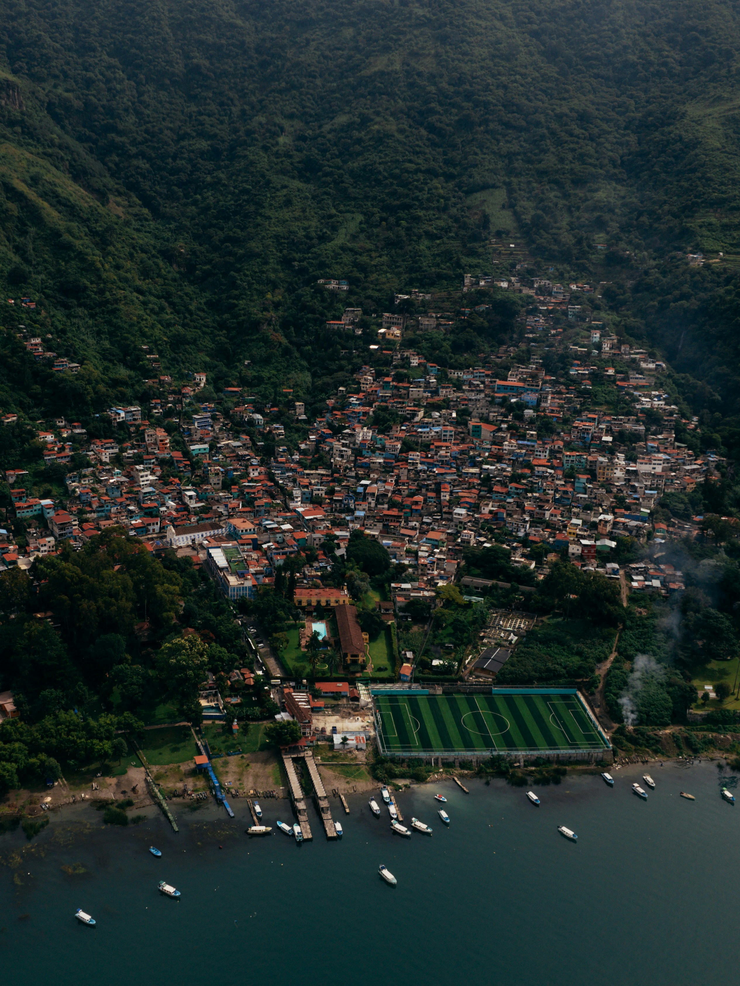 Ribbed valley town by a moored harbor: aerial view of a dense riverside settlement with a football pitch