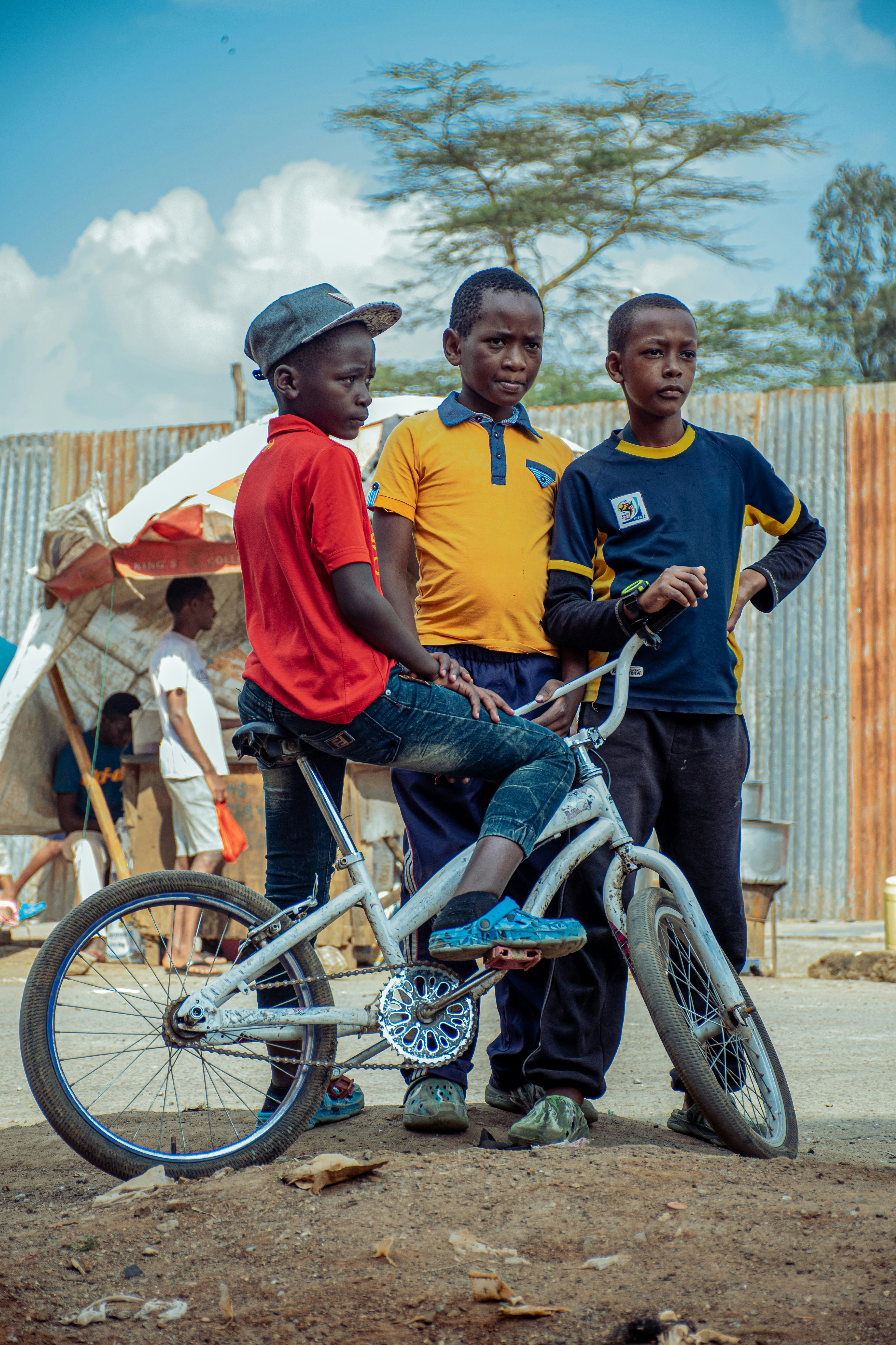 Three children standing together, one perched on a white bicycle, set against a backdrop of urban life with a colorful tent.