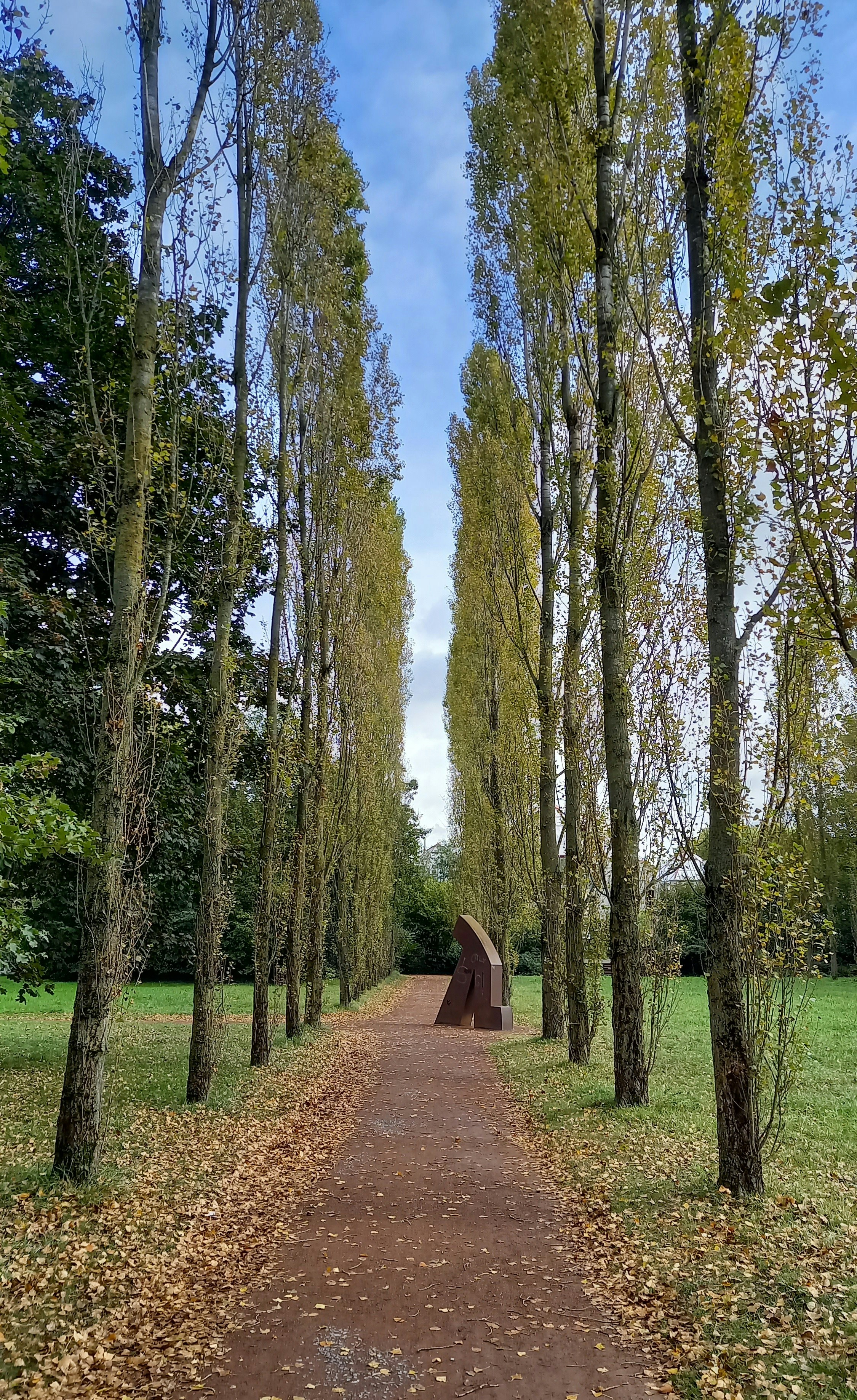 Dirt path in a park is framed by tall, leaf-shedding trees, leading to a rust-colored abstract sculpture in the distance.