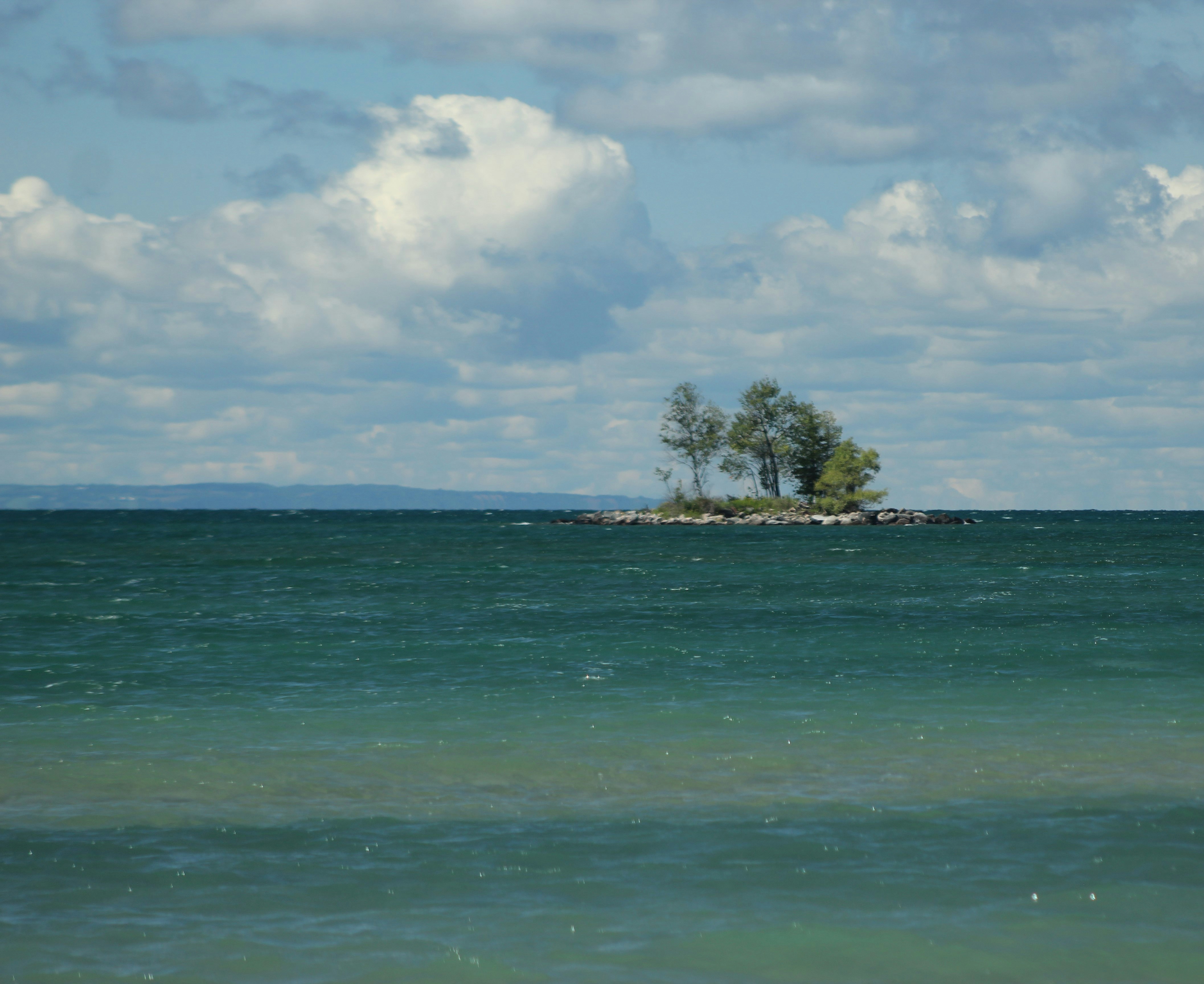 Tiny island with a few trees surrounded by expansive ocean under a partly cloudy sky.