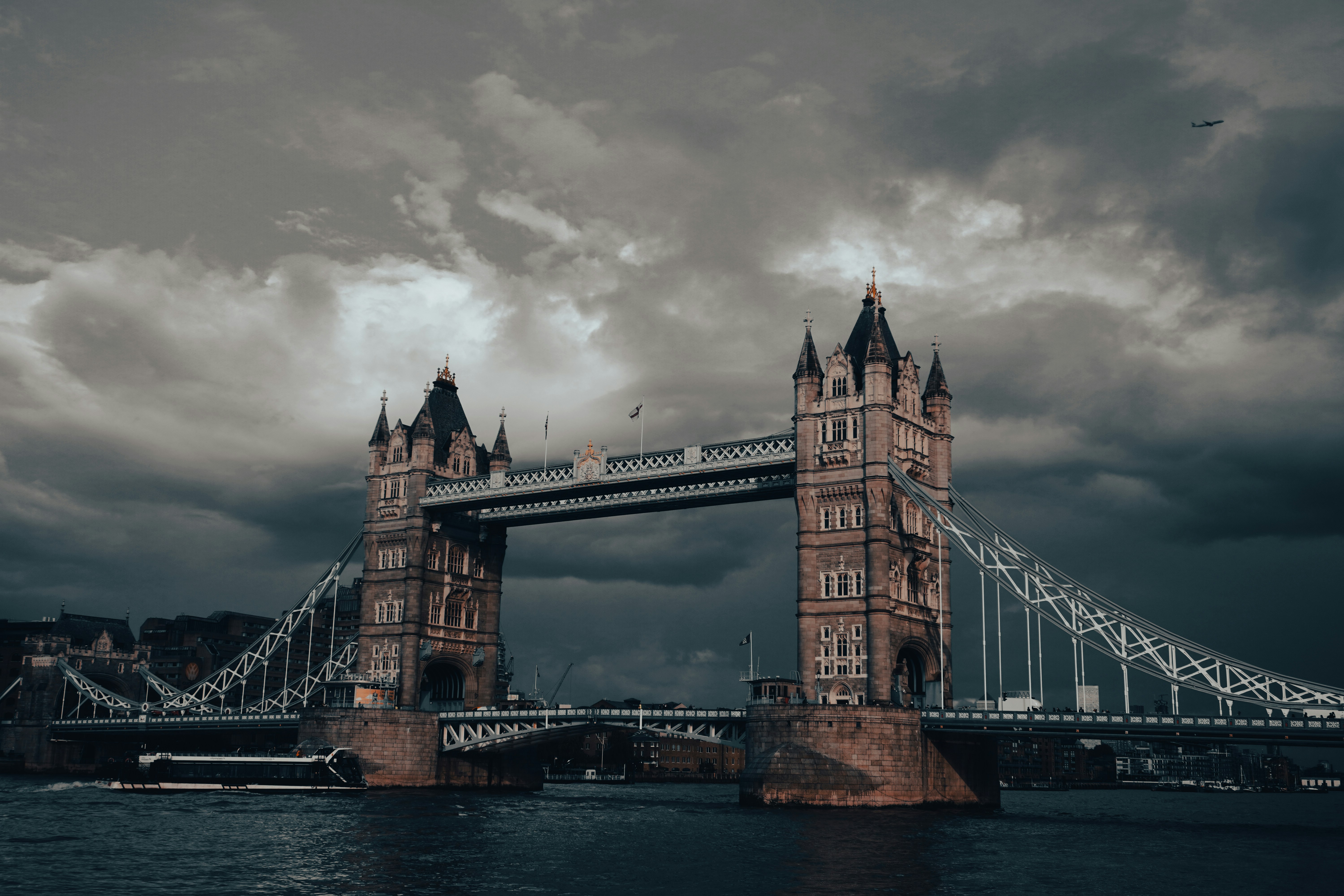 Tower Bridge stands majestically against a moody sky, showcasing its intricate architecture and iconic towers. The scene captures a dramatic interplay of light and shadow.