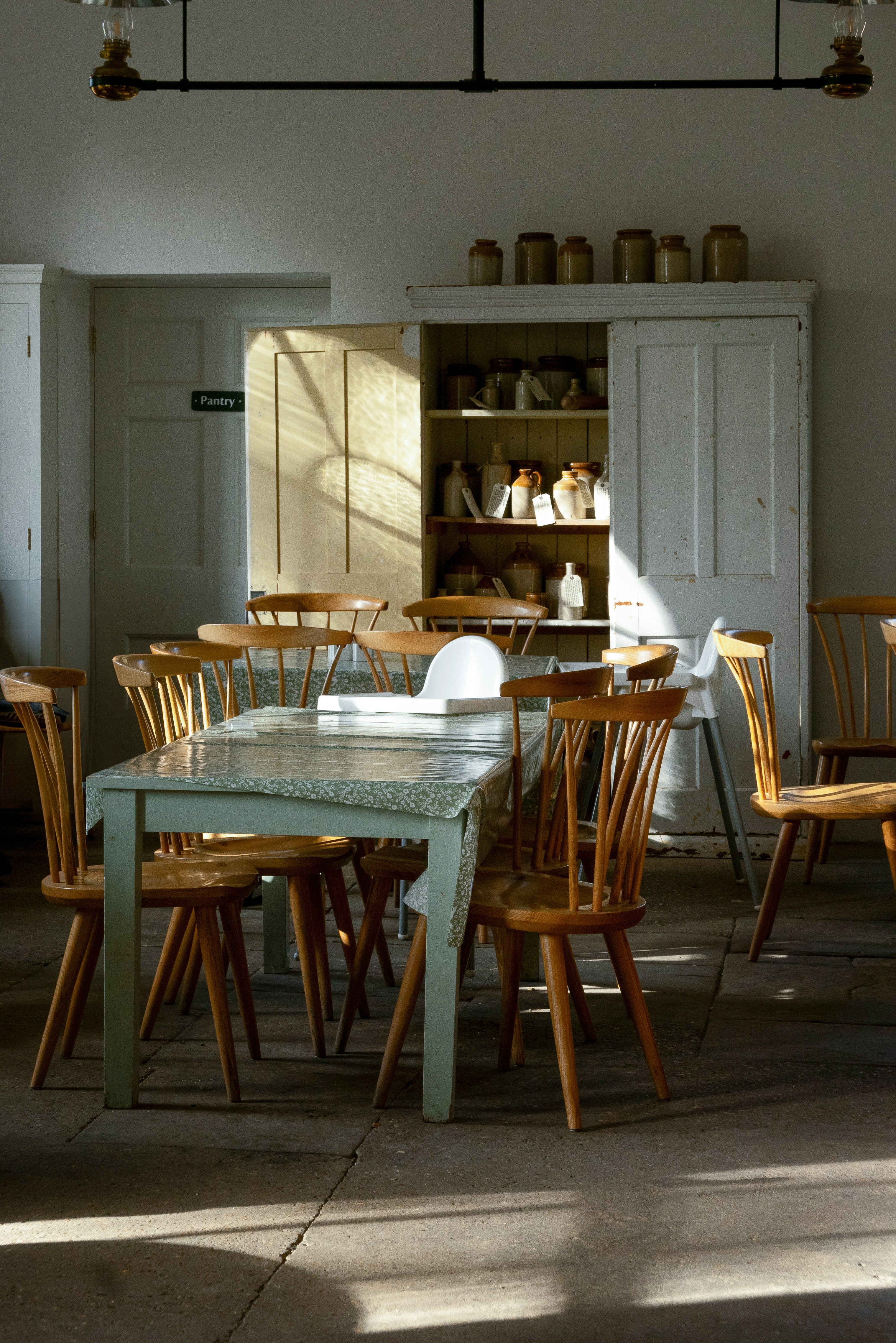 Sunlight casts long shadows across a rustic dining room with wooden chairs and a vintage cupboard.