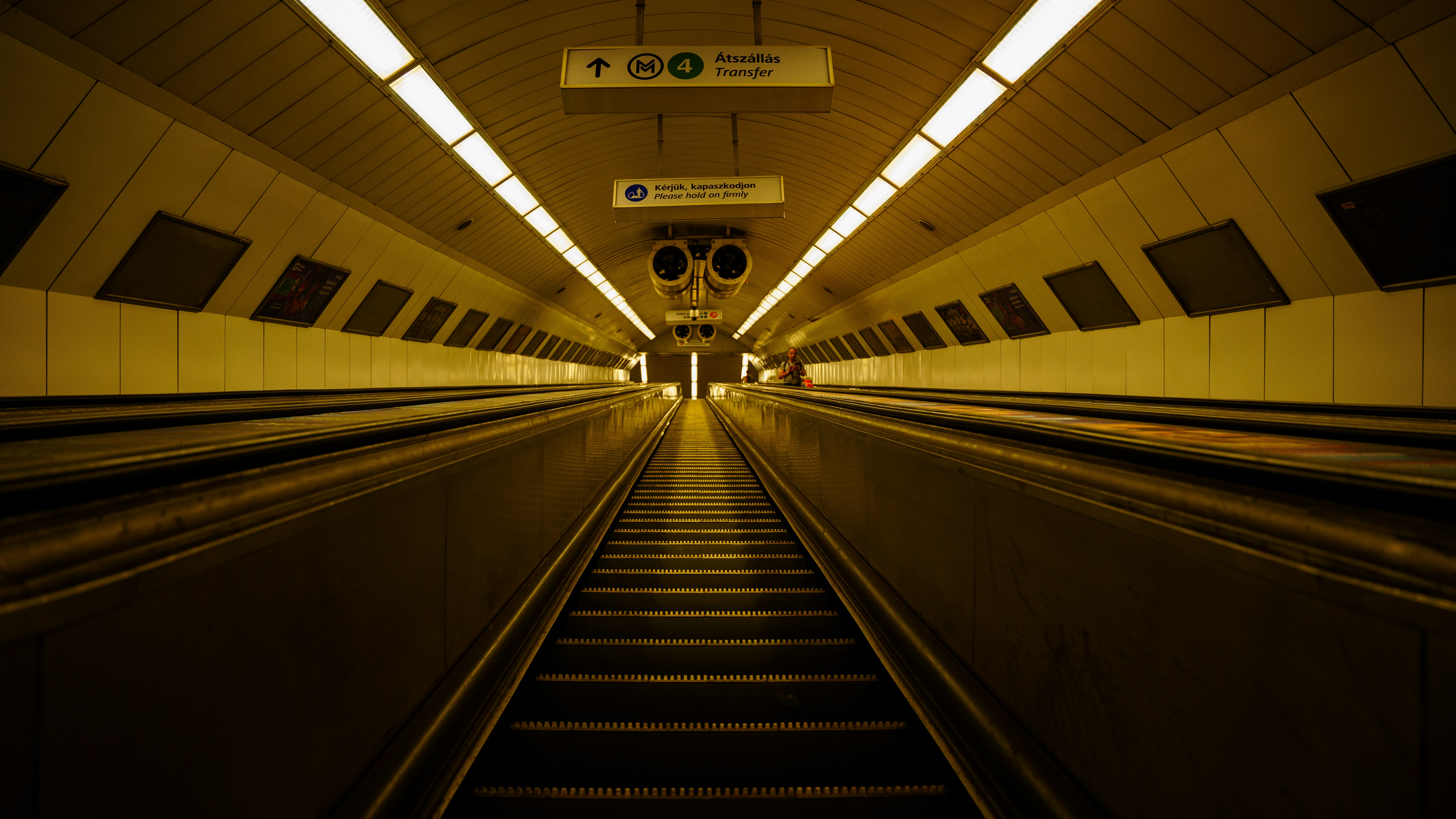 Escalator leading down into a subway station, illuminated by overhead lights and flanked by signage. The perspective creates a sense of depth and movement.