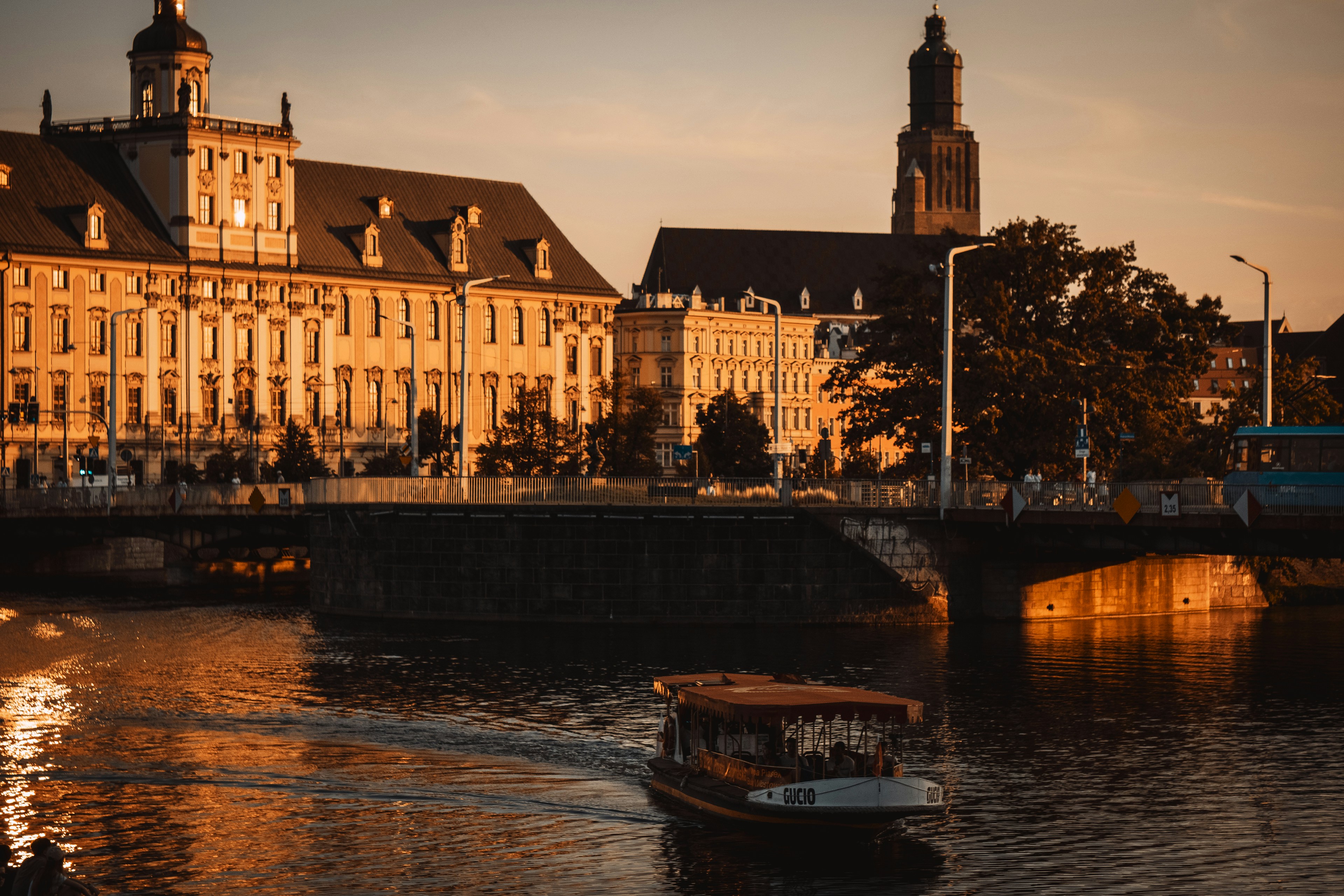 A charming riverboat glides past historic architecture bathed in warm golden light during sunset.