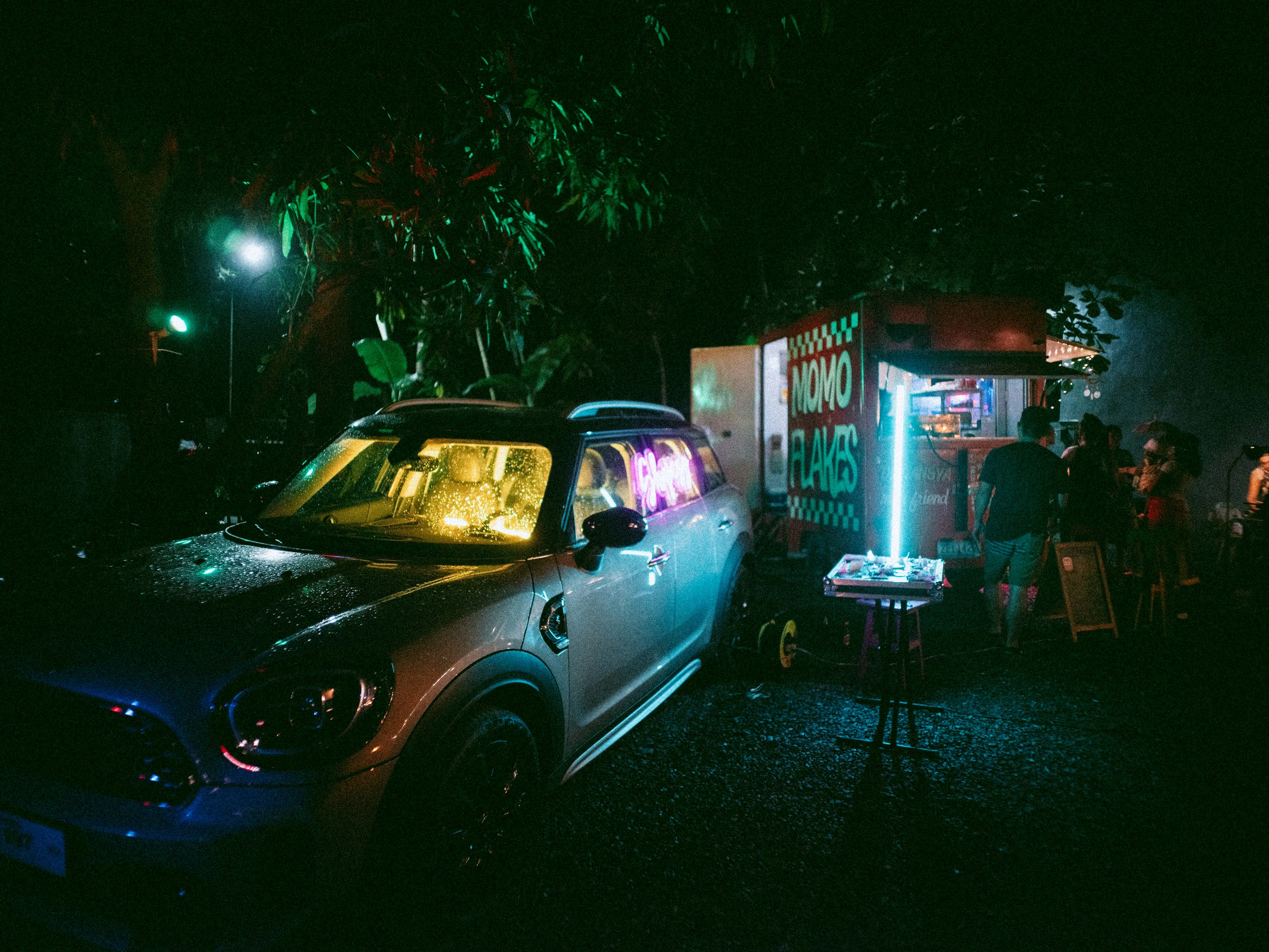 Nighttime photograph of a neon-lit car parked beside a street food stall, with people gathering under colorful lights.