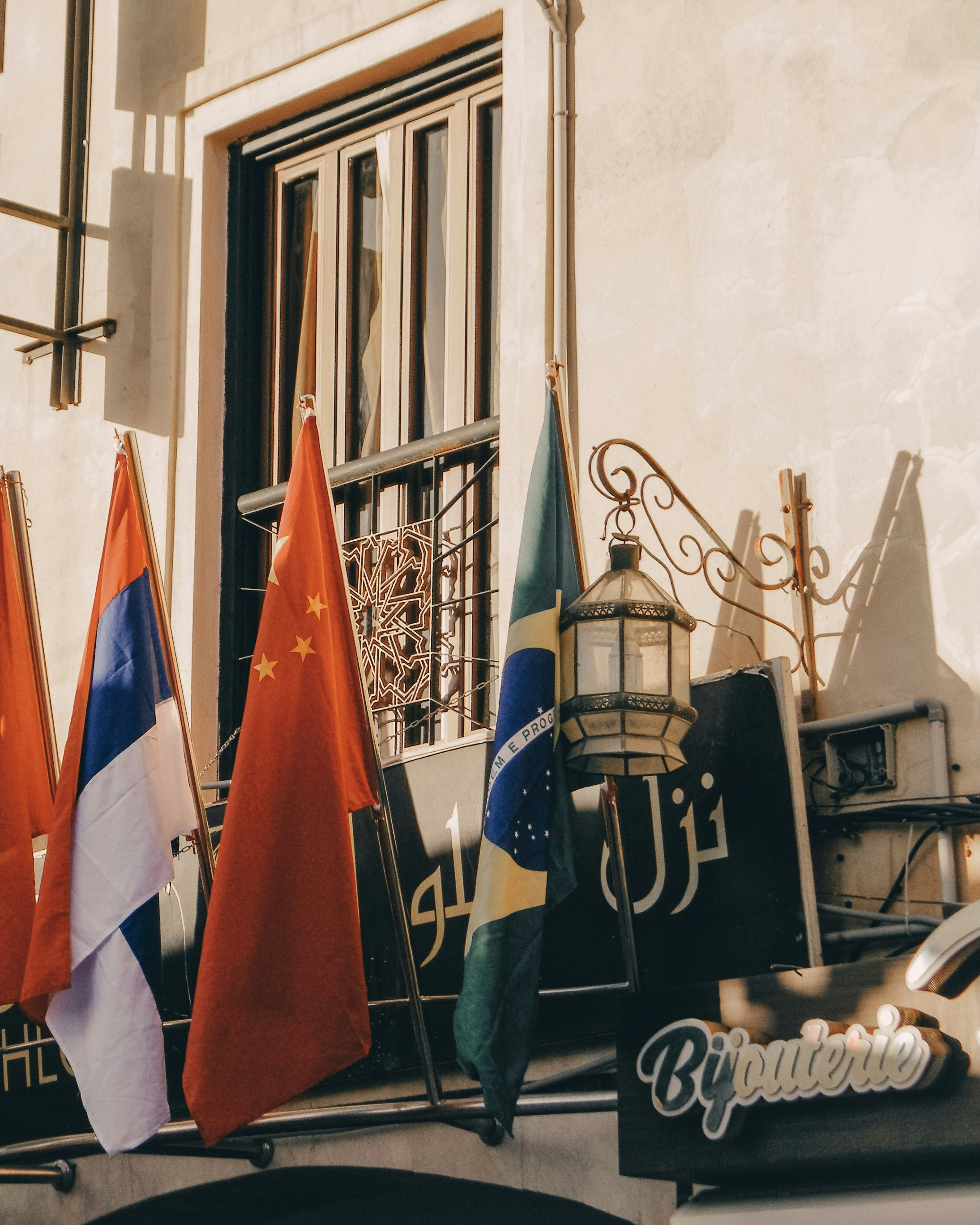 Photograph of a sunlit storefront corner with flags fluttering in front of a window and a decorative lantern beside a jewelry sign. The scene emphasizes flag textures and architectural details.