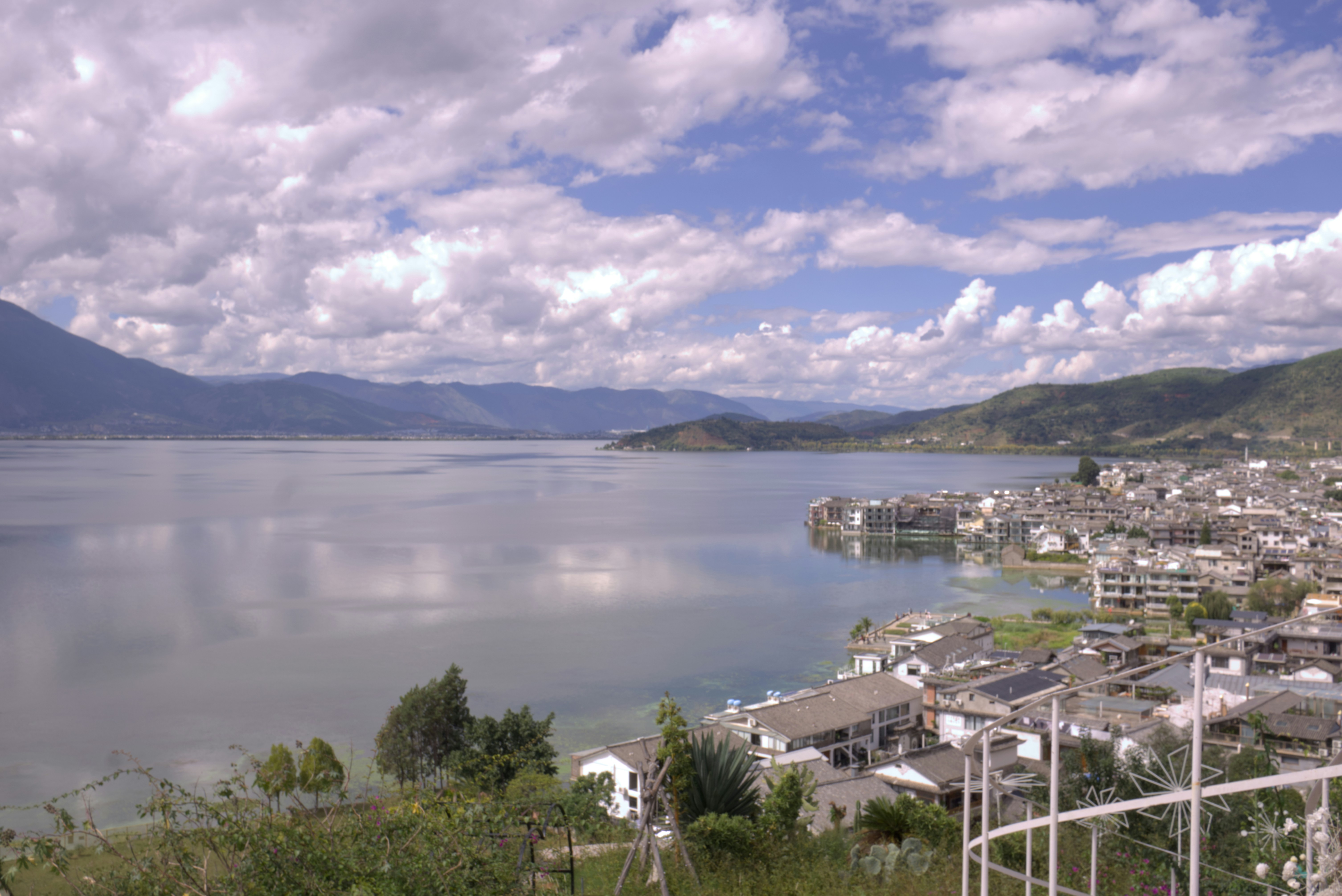 Serene waters of a lake bordered by lush hills under a sky filled with fluffy clouds.