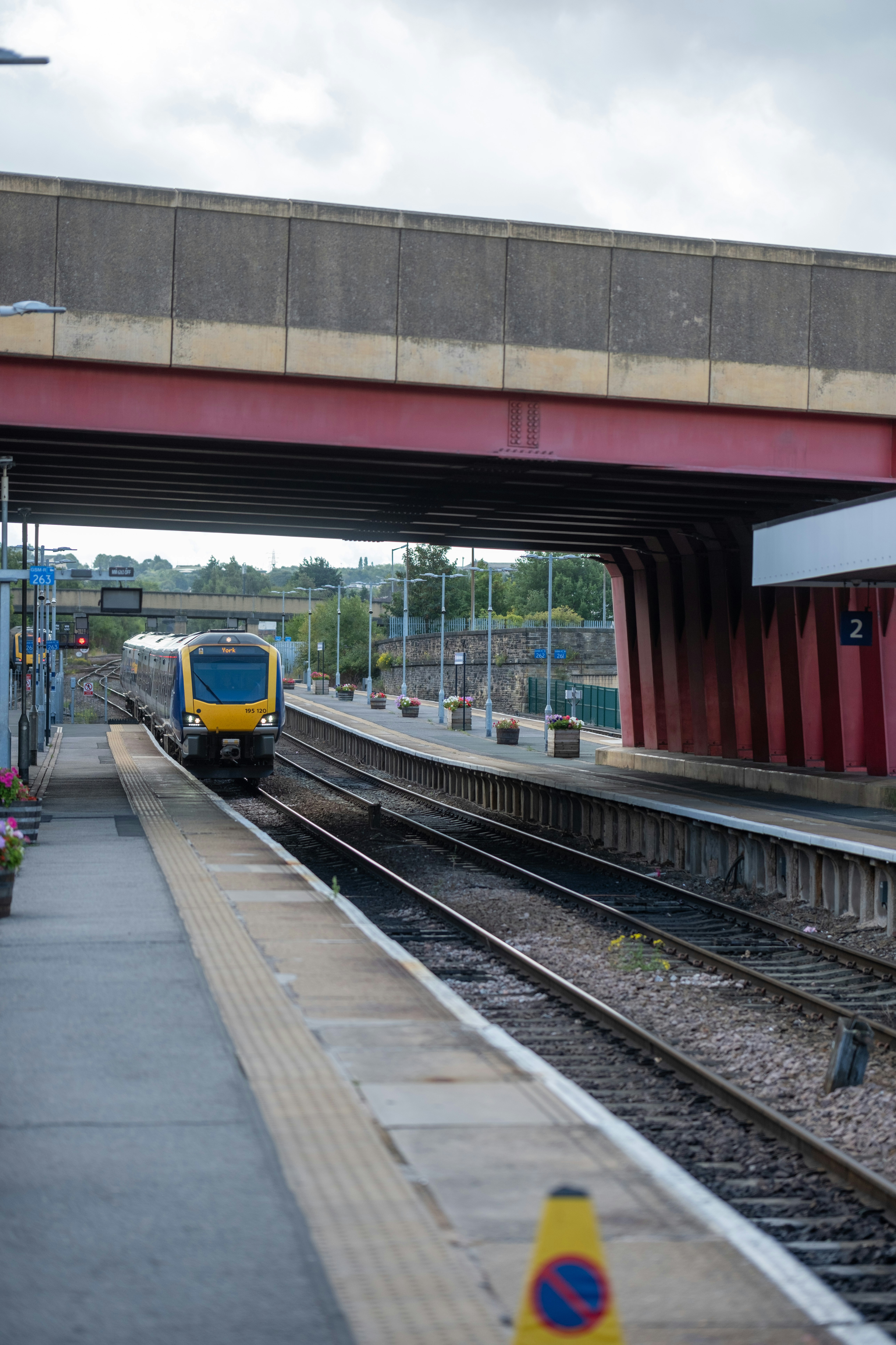 Train approaching a station platform under a red bridge on an overcast day.