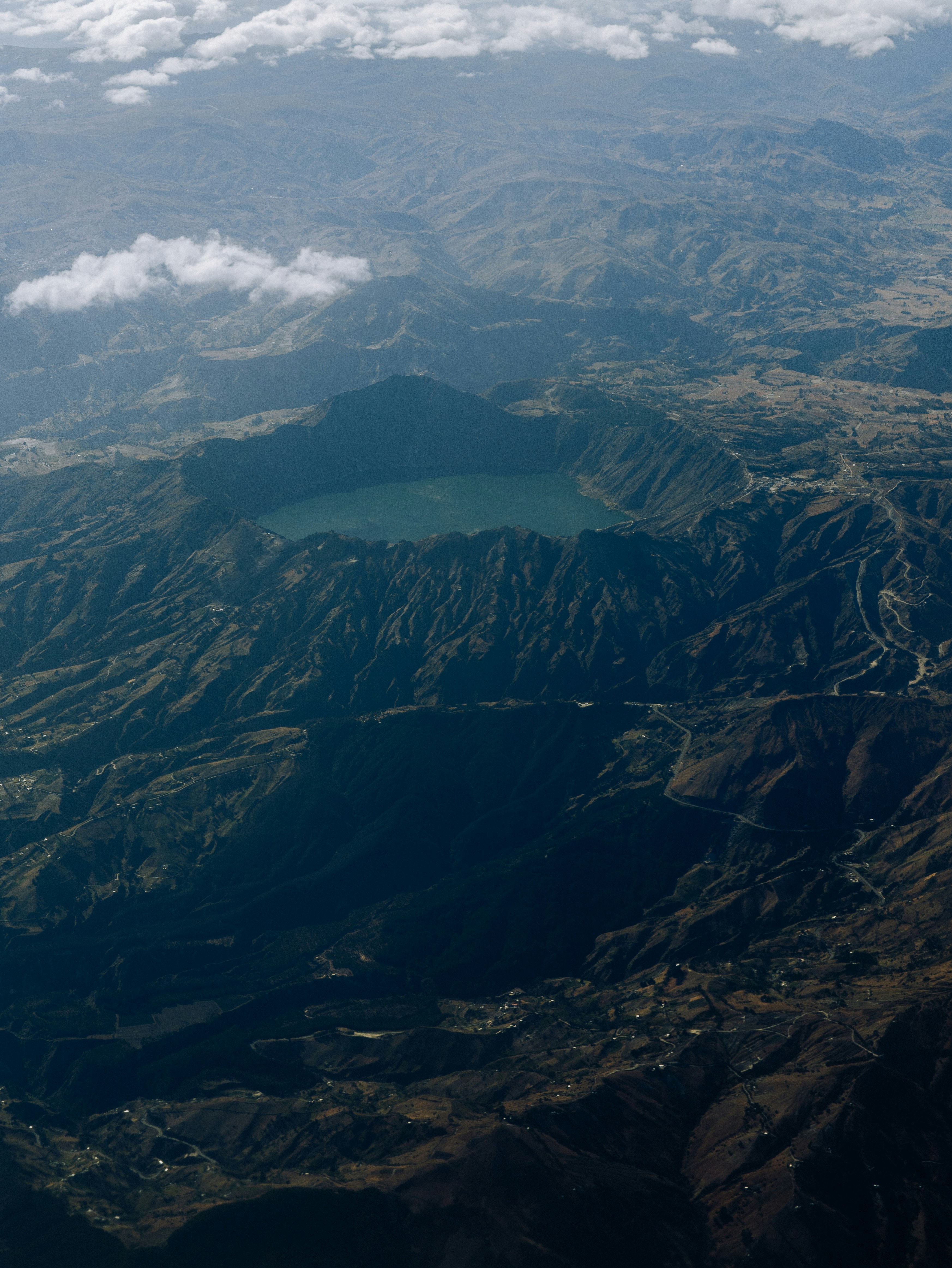 Aerial view showcasing a tranquil crater lake surrounded by rugged mountains and valleys, with wisps of clouds above. 
