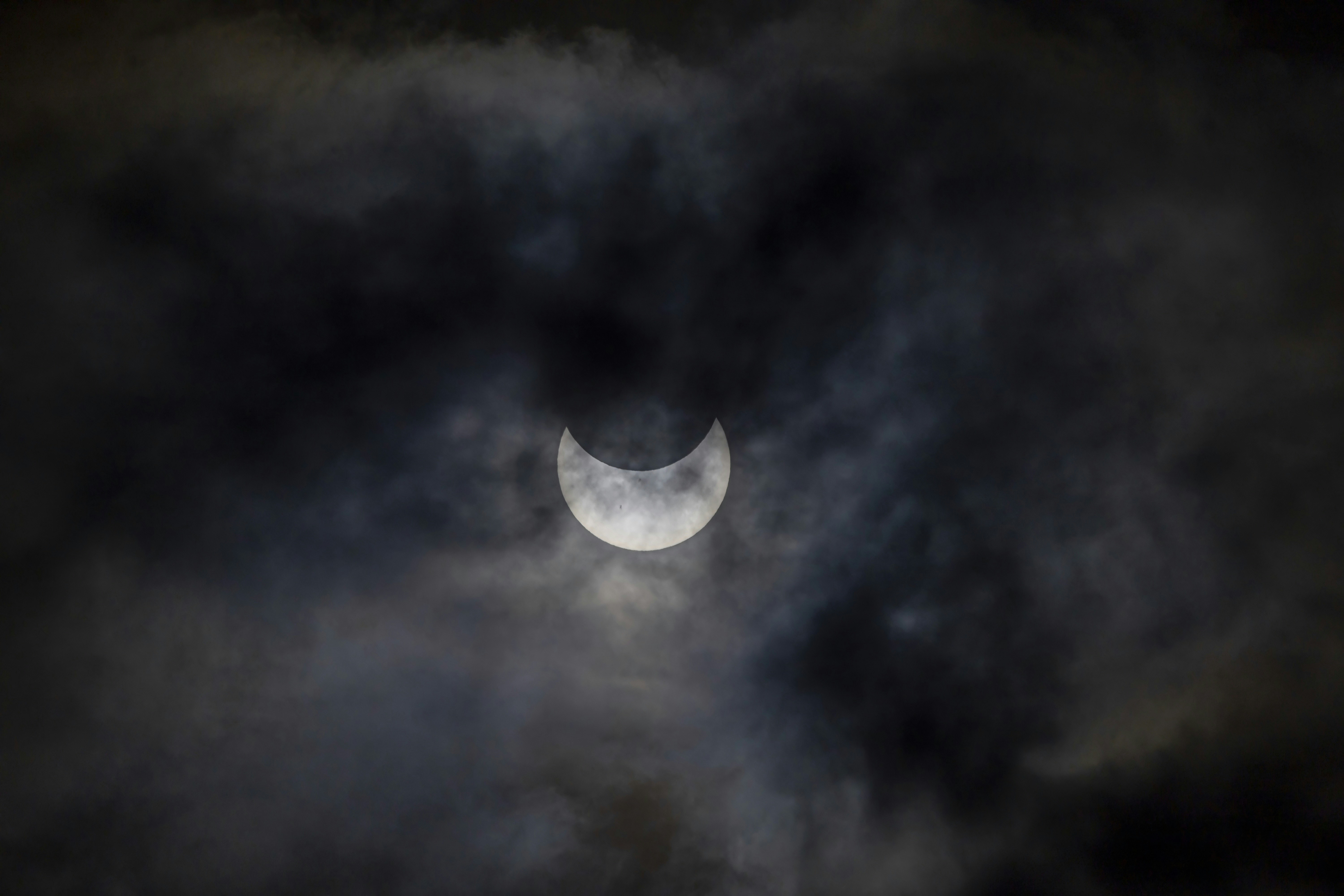 A photograph capturing a partial solar eclipse glimpsed through a stormy veil of clouds, with a pale crescent sun peeking through.