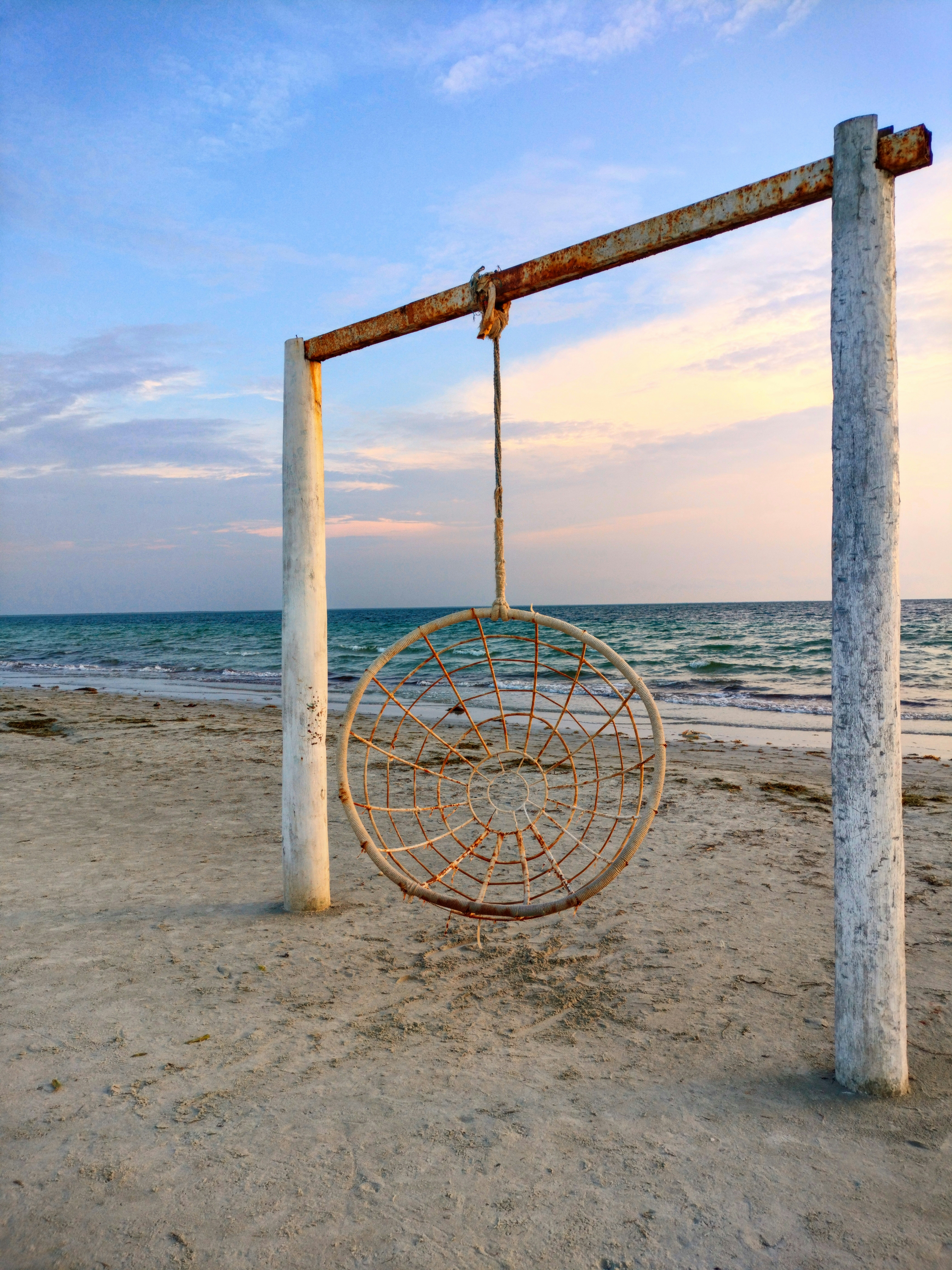 Circular rope swing suspended between weathered white posts on a sandy beach, with the calm sea and pastel sunset filling the horizon.