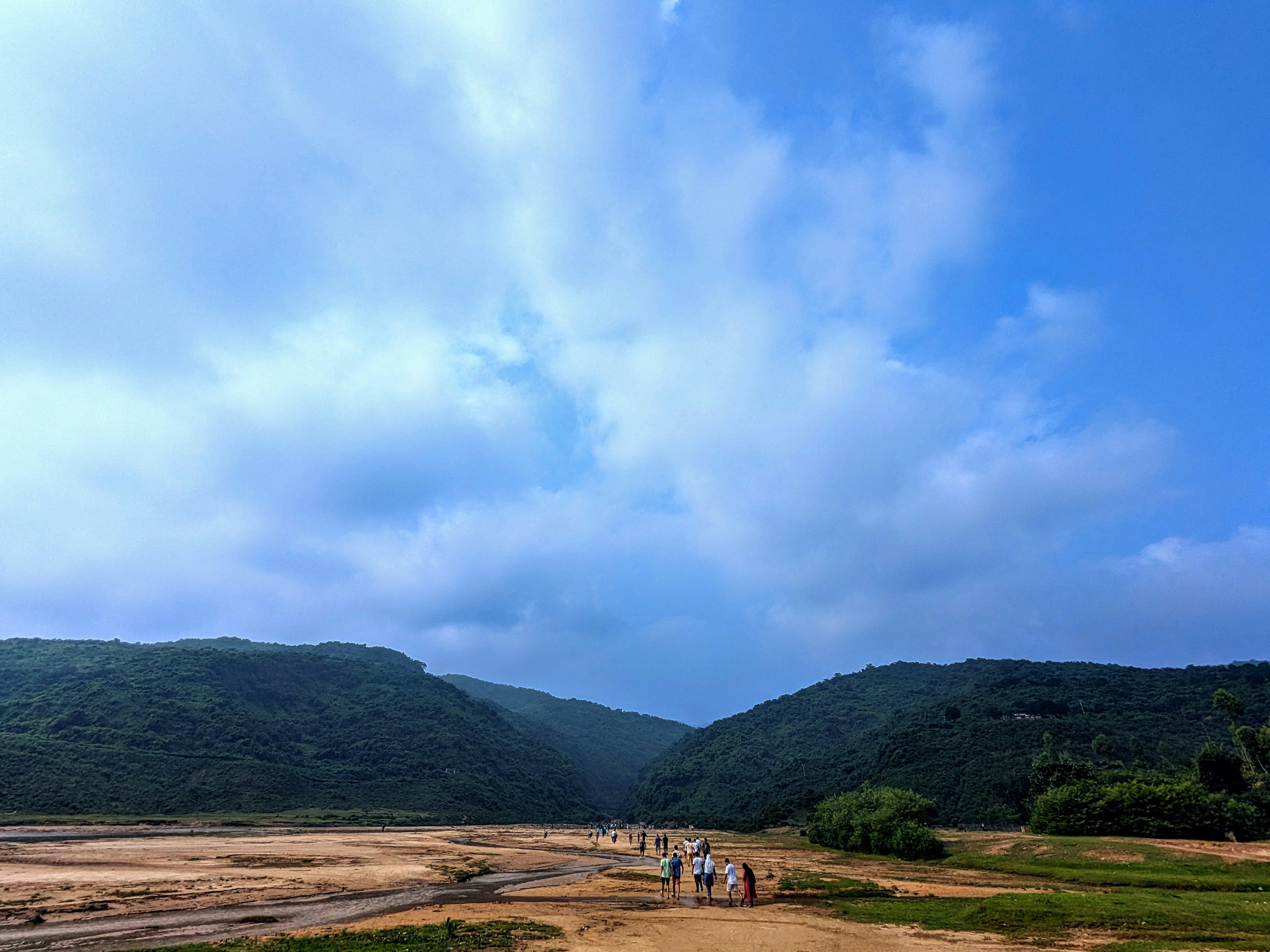Hikers on a quiet dry riverbed framed by forested hills under a cloud-dappled blue sky