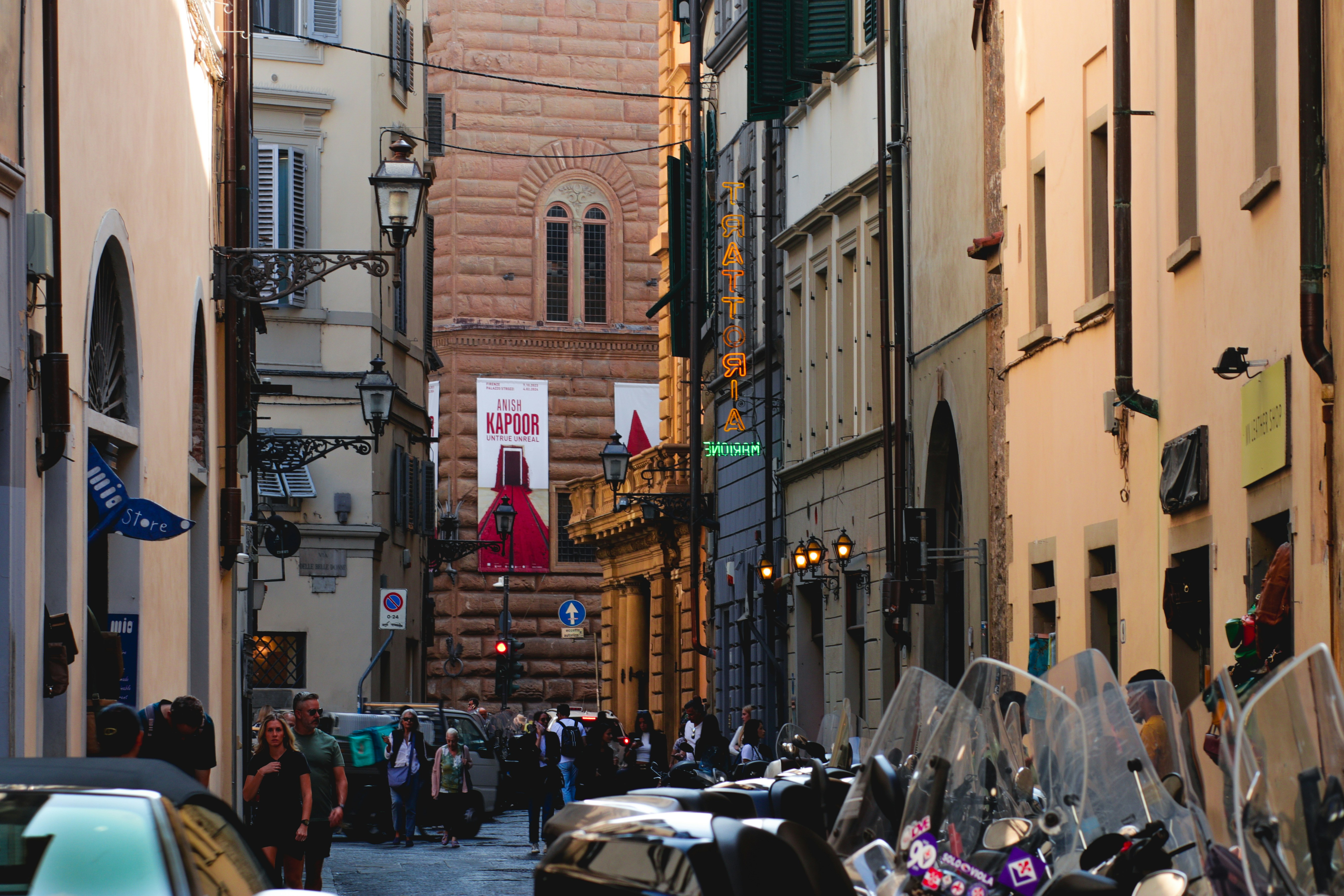 Narrow city street bustling with pedestrians and lined with parked motorcycles, featuring a prominent advertisement for Kapoor on a building façade.