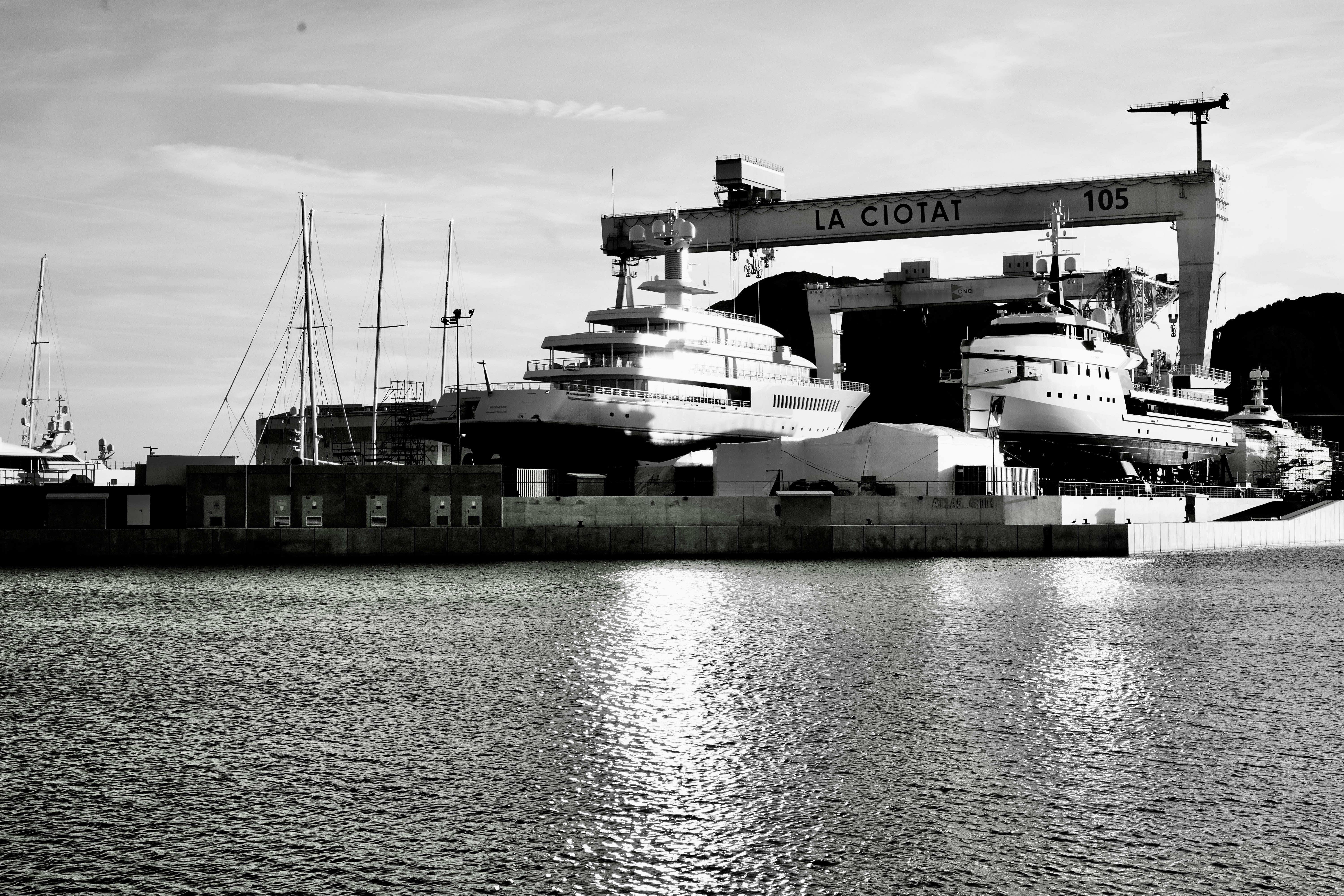 Black and white view of a shipyard with large vessels under construction, framed by a crane and calm waters.