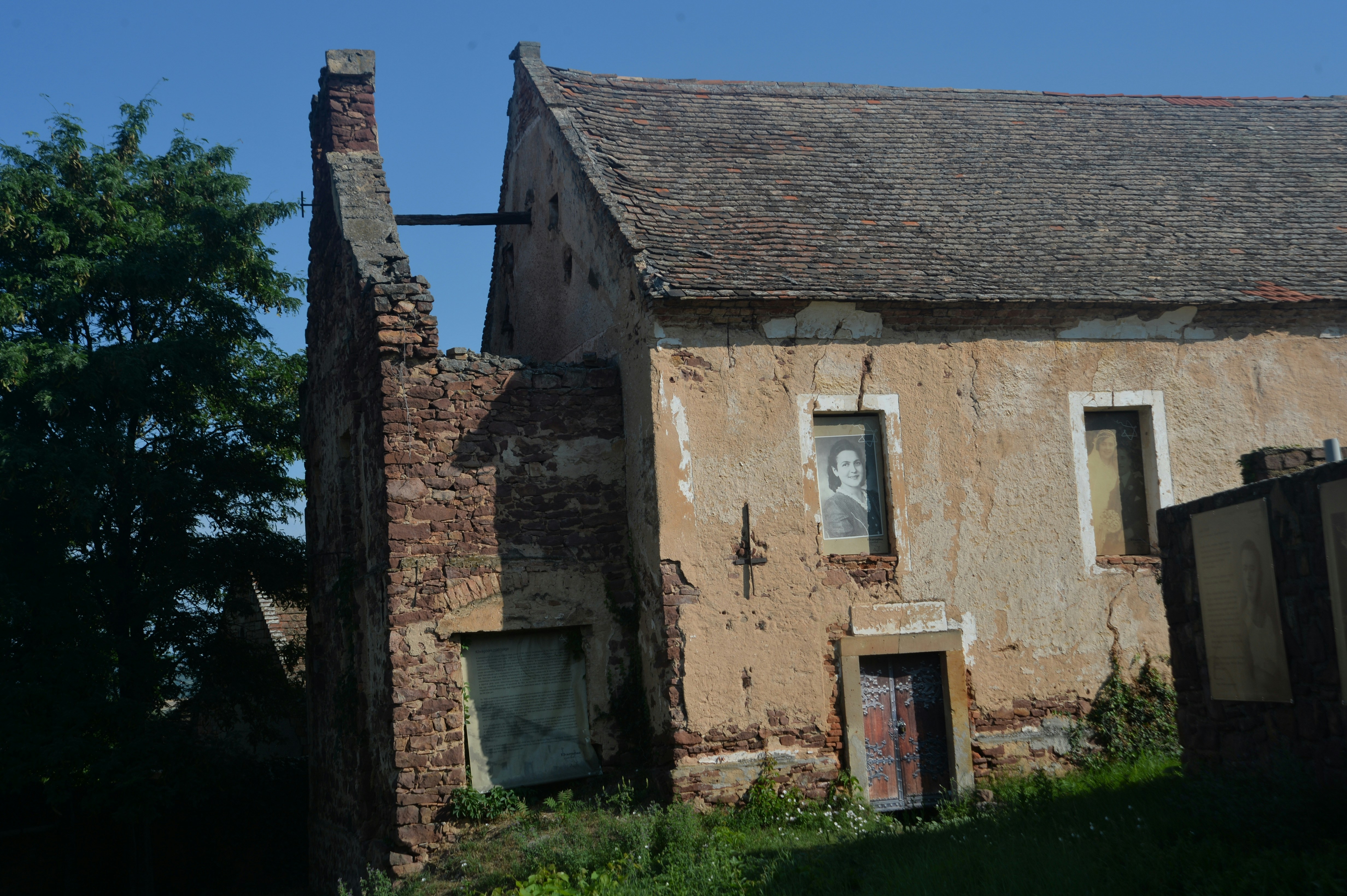 Dilapidated building with crumbling walls and a ghostly portrait visible through a window, surrounded by overgrown greenery.