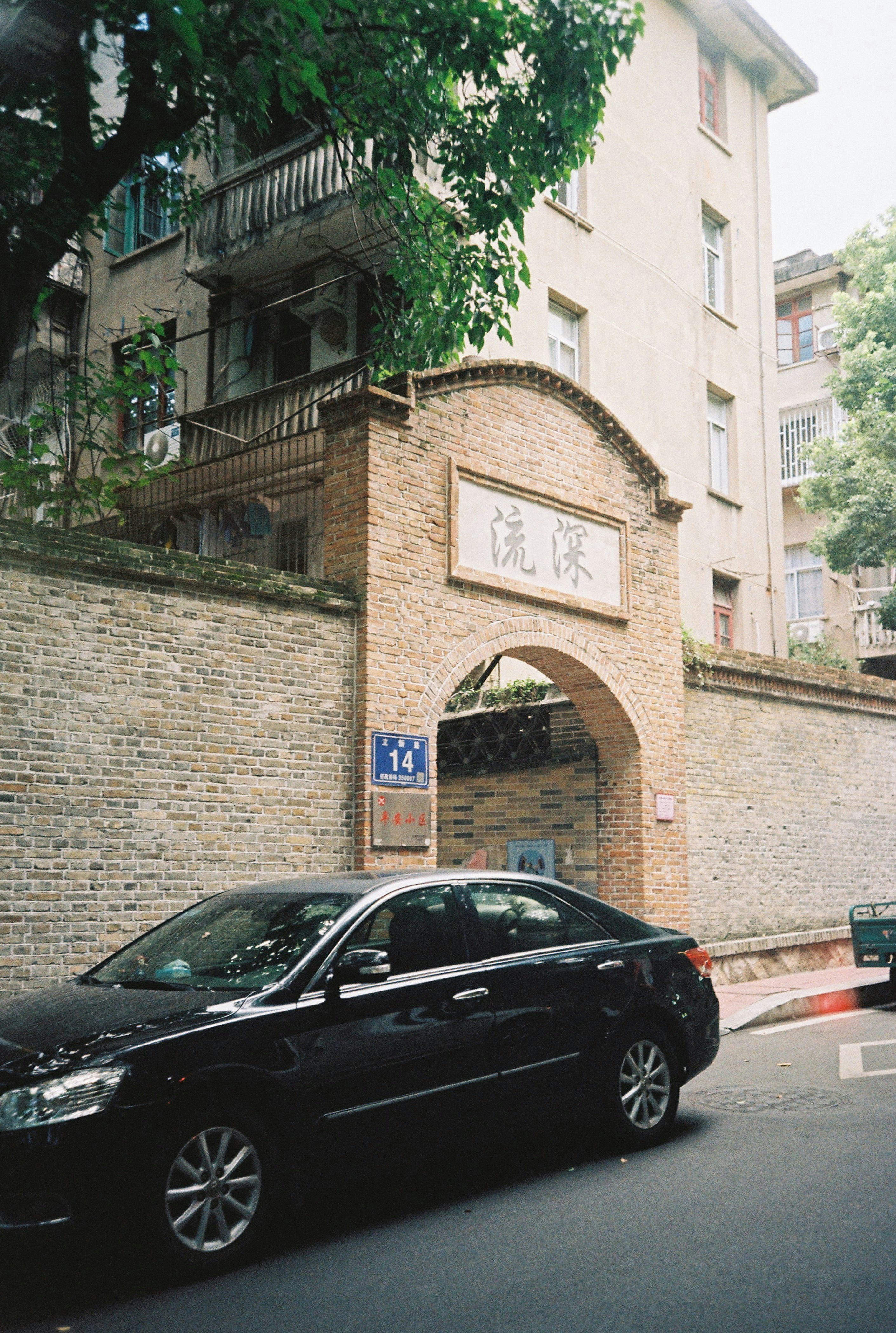 Archway of the Quiet Block: Brick Gate, Leafy Canopy, and a Passing Sedan