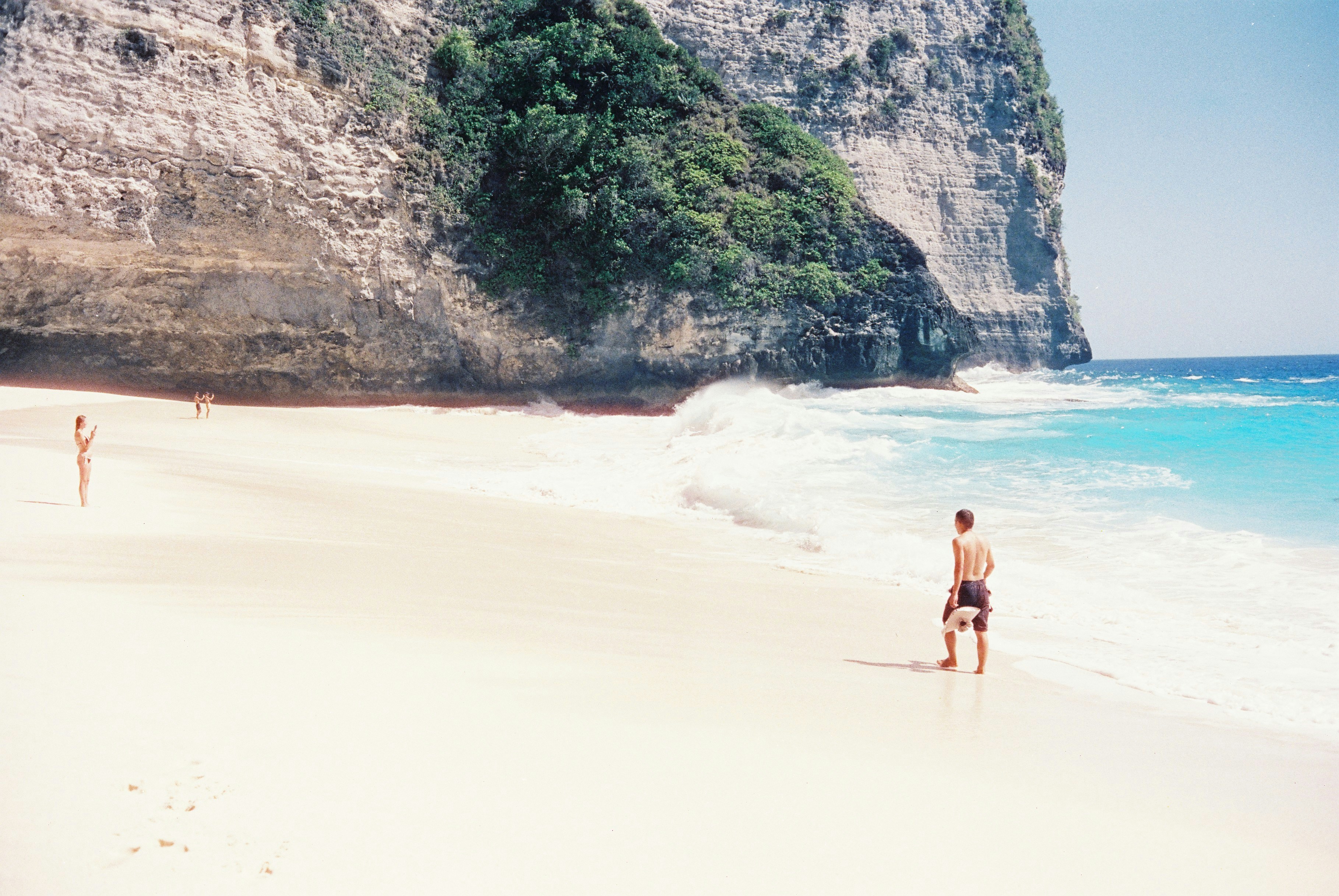 Bright beach scene with pale sand, turquoise waves, and a cliff on the left; a lone swimmer stands near the waterline, while a small figure appears farther left.