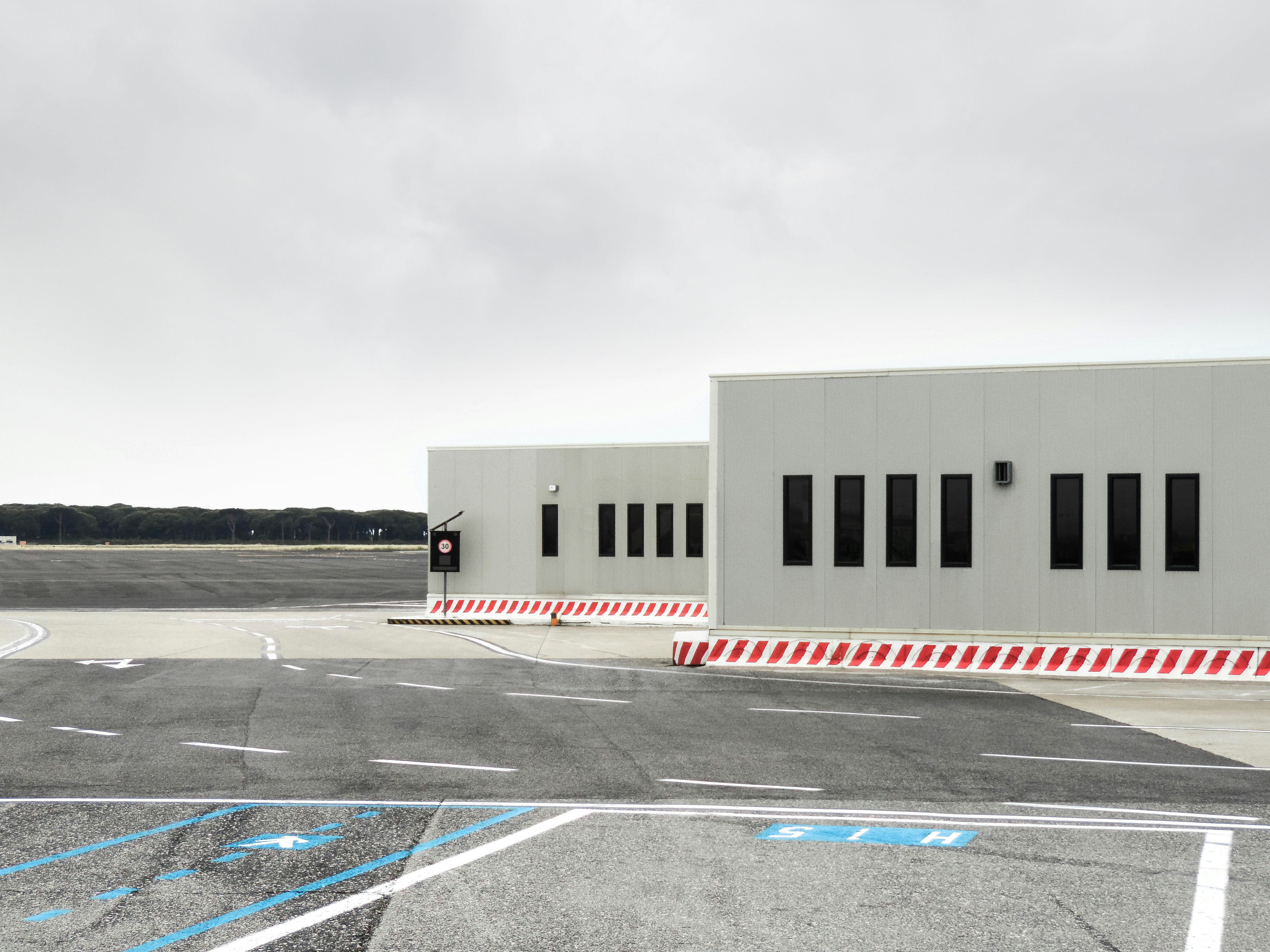 Photograph: Wide, empty tarmac leads to a white, boxy hangar with red-striped barriers under a gray, overcast sky.