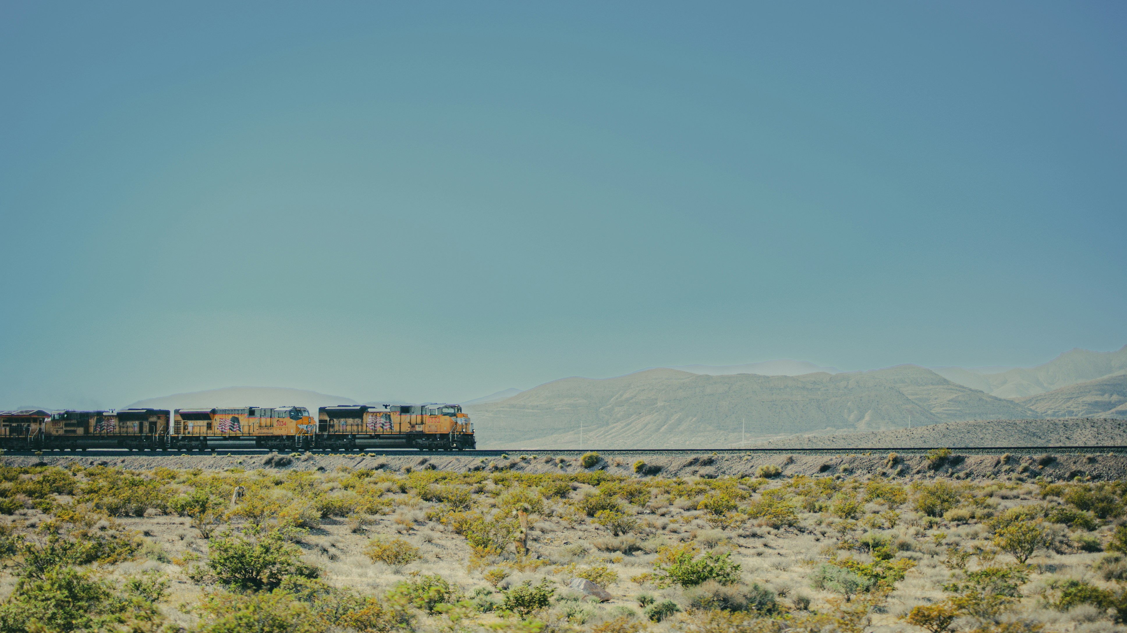 Freight train traversing a vast, arid landscape under a clear blue sky, highlighting the contrast between machinery and nature.