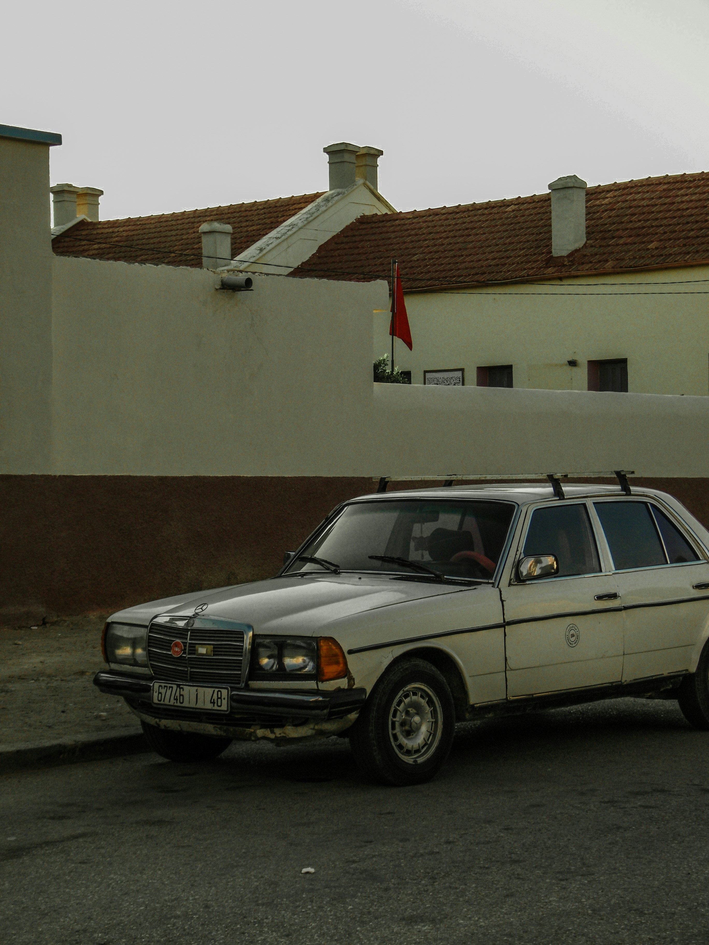 Vintage Mercedes sedan parked along a sunlit walled street, with a red flag raised near a pale building in the background.