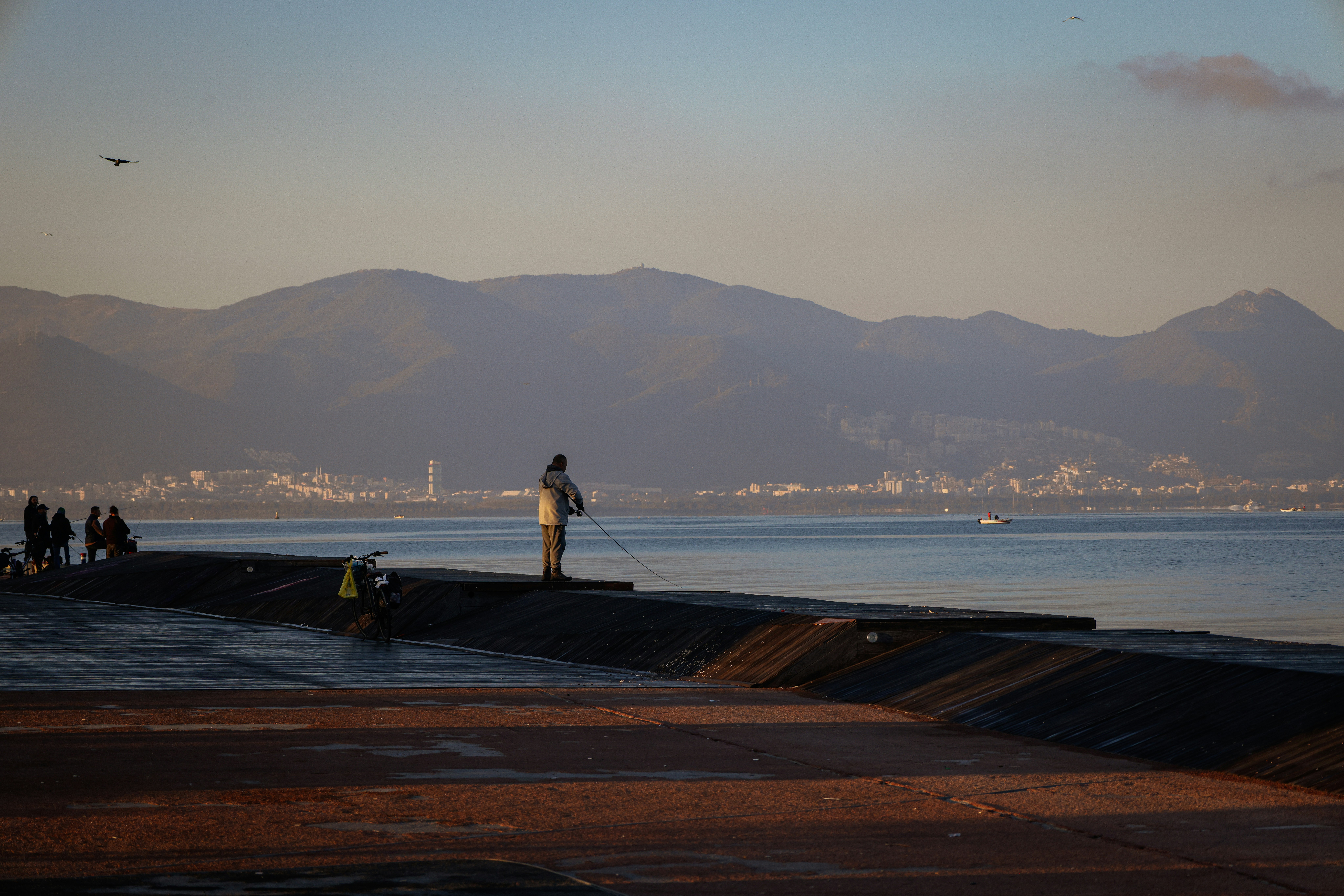 Individuals stand on a pier overlooking calm waters with distant mountains under a clear sky.