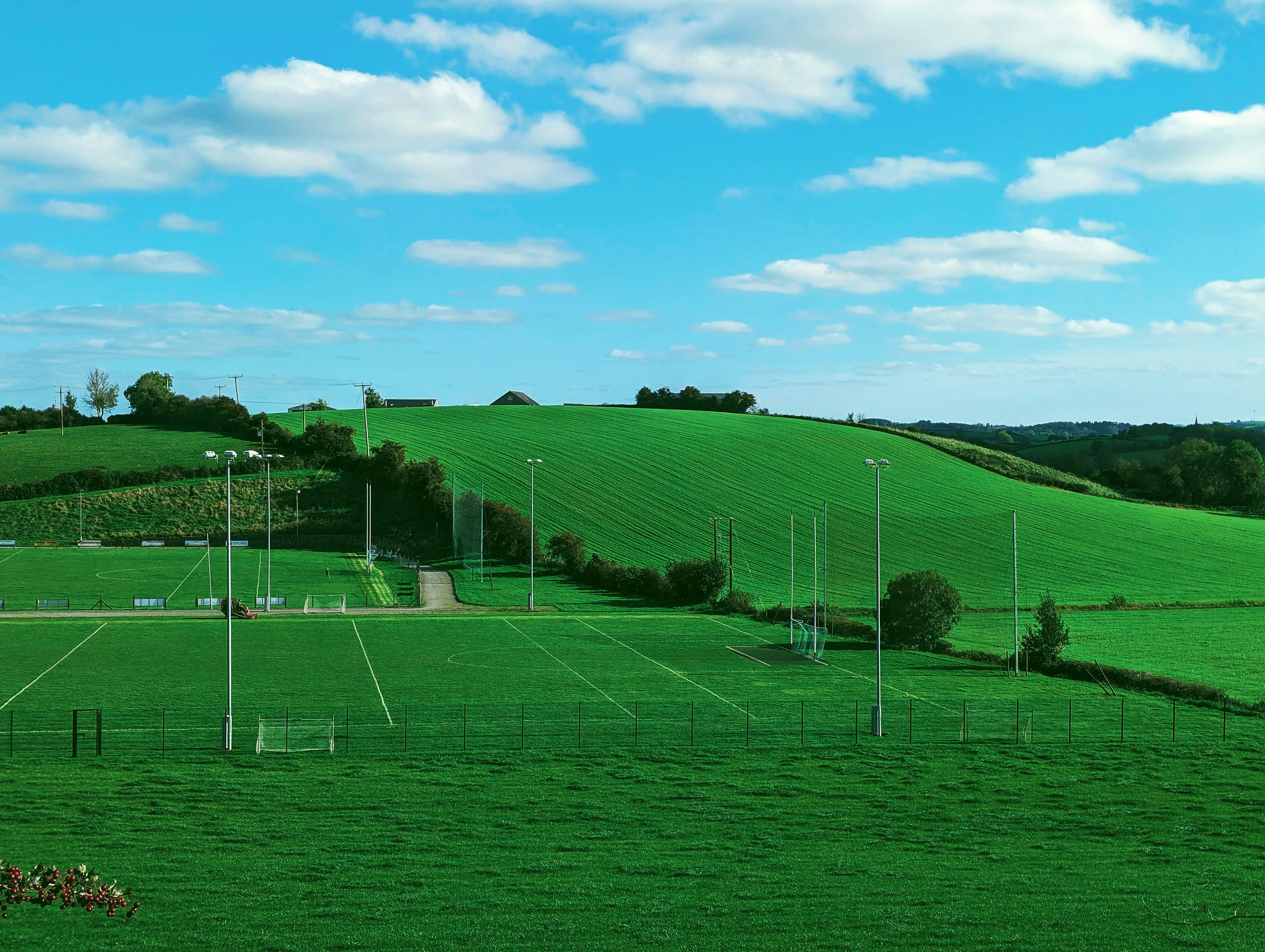 Lush green fields frame a soccer pitch with goalposts beneath a clear blue sky. Rolling countryside provides a calm, expansive backdrop.