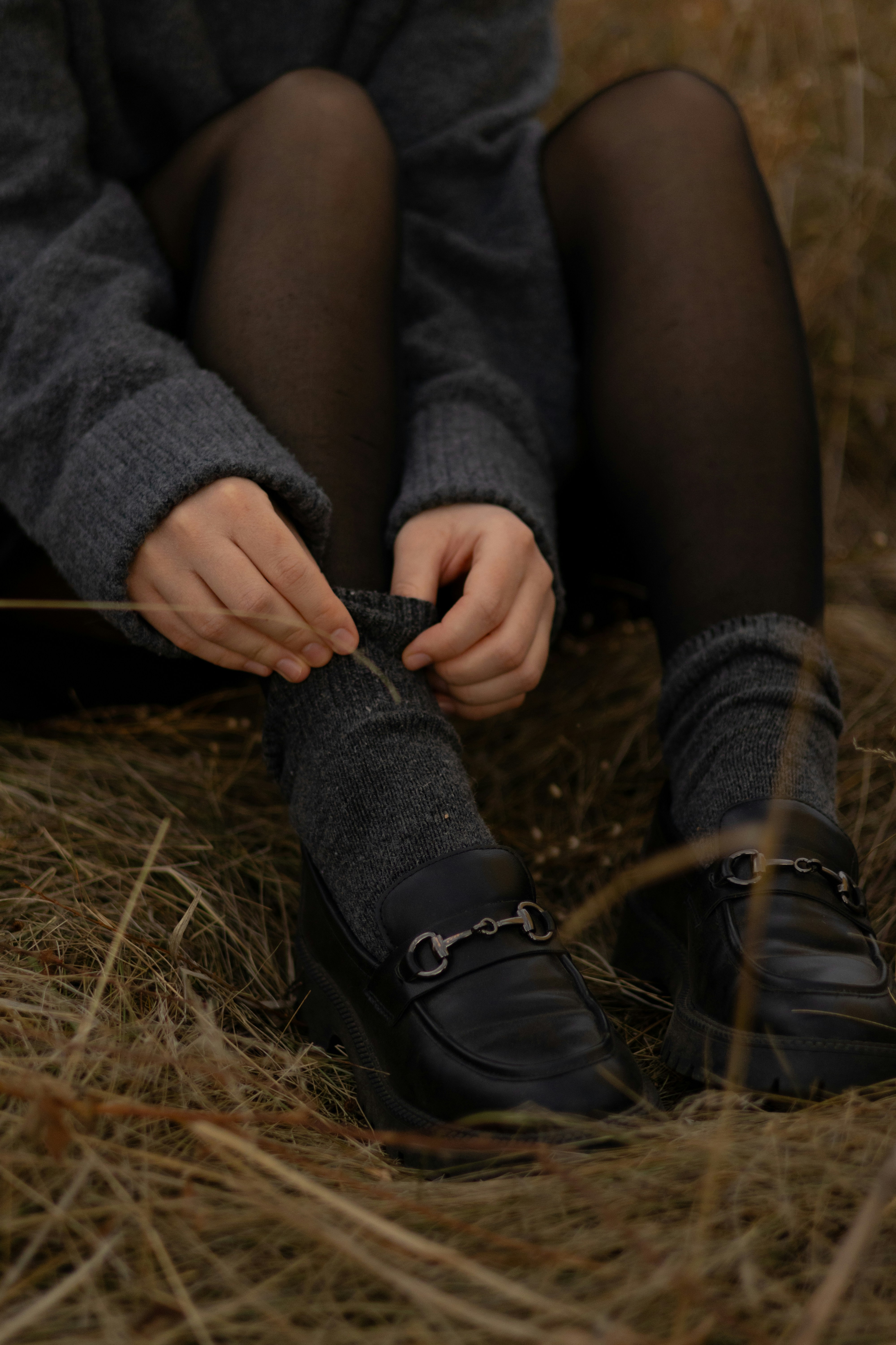 Close-up candid photograph of hands adjusting thick wool socks over loafers, seated amid dry grass in muted autumn tones.