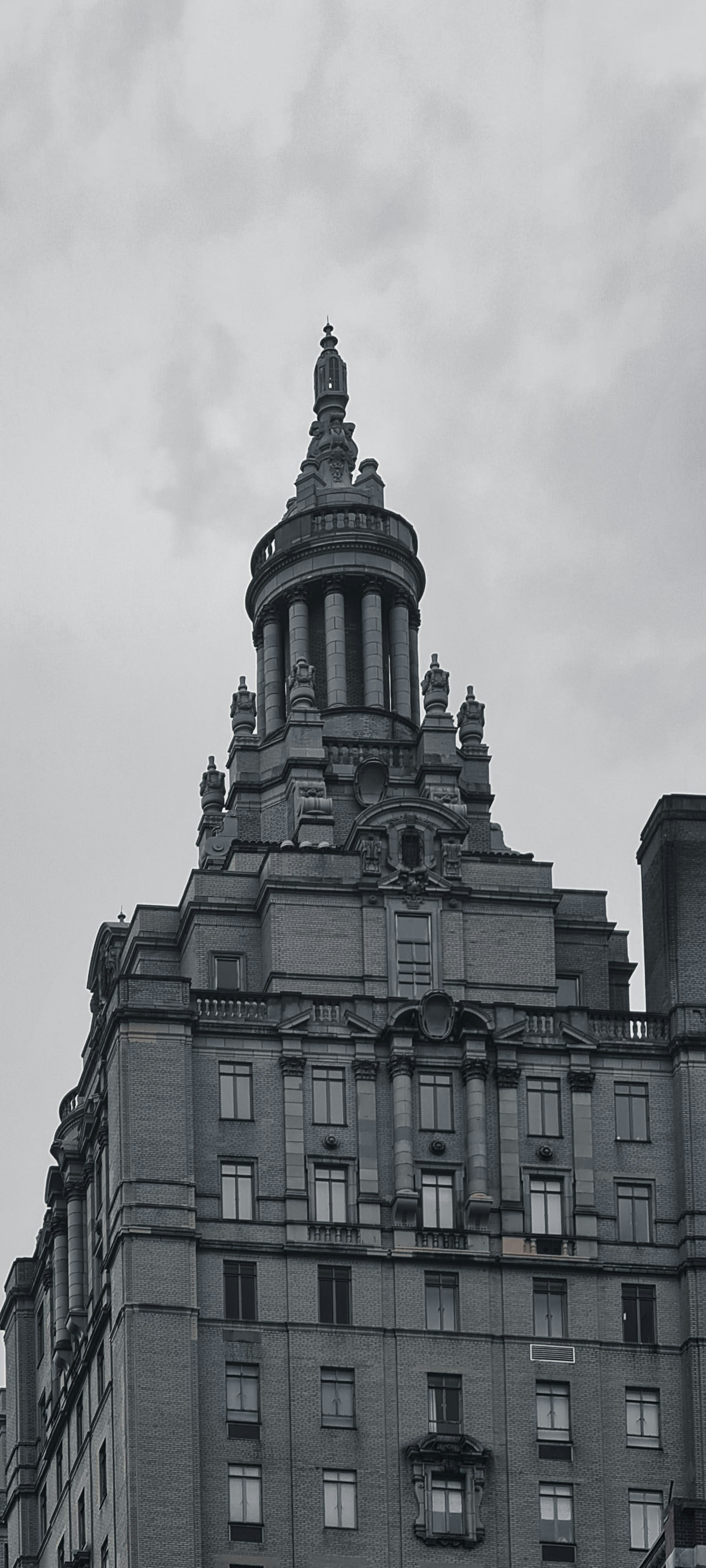 Grayscale photograph of a tall, ornate brick tower rising above the lower facade against a cloudy sky. The shot emphasizes architectural detail and vertical scale.