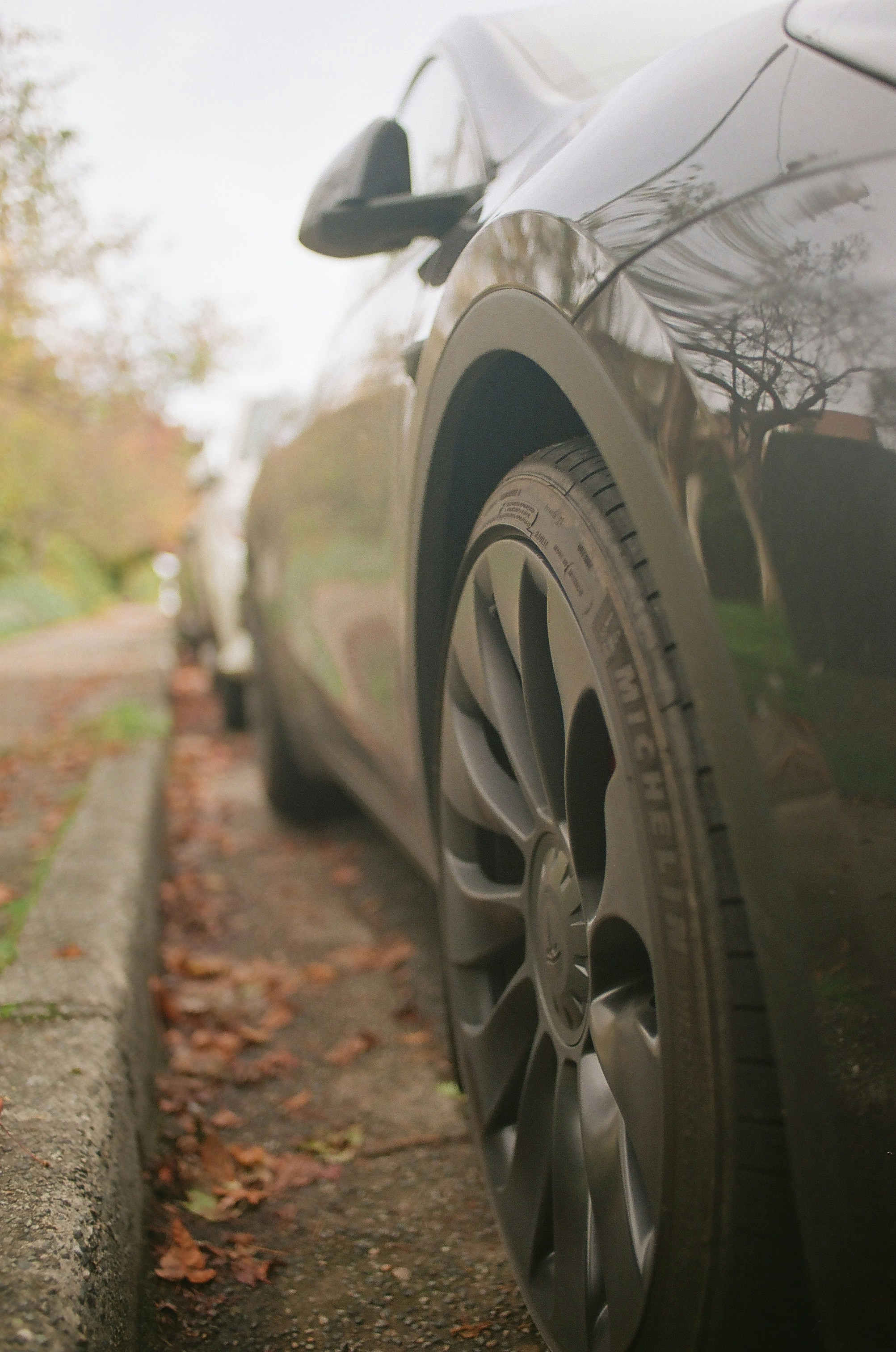 Close-up photograph of a glossy car wheel along a leaf-strewn curb with soft background bokeh.