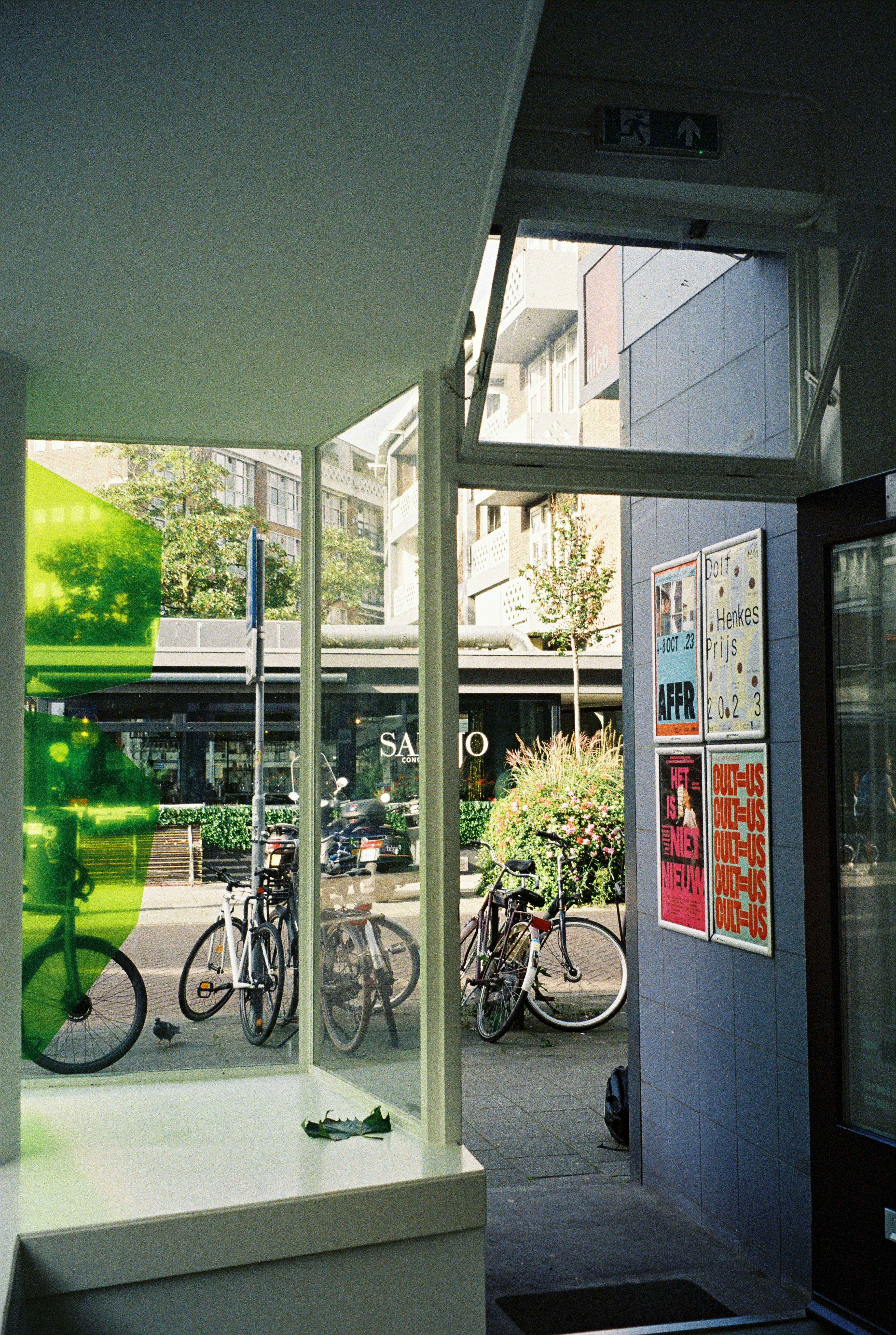 Urban storefront scene viewed through a display window, with bicycles parked along the curb and a lime-green pane on the left. Posters color the blue wall to the right.