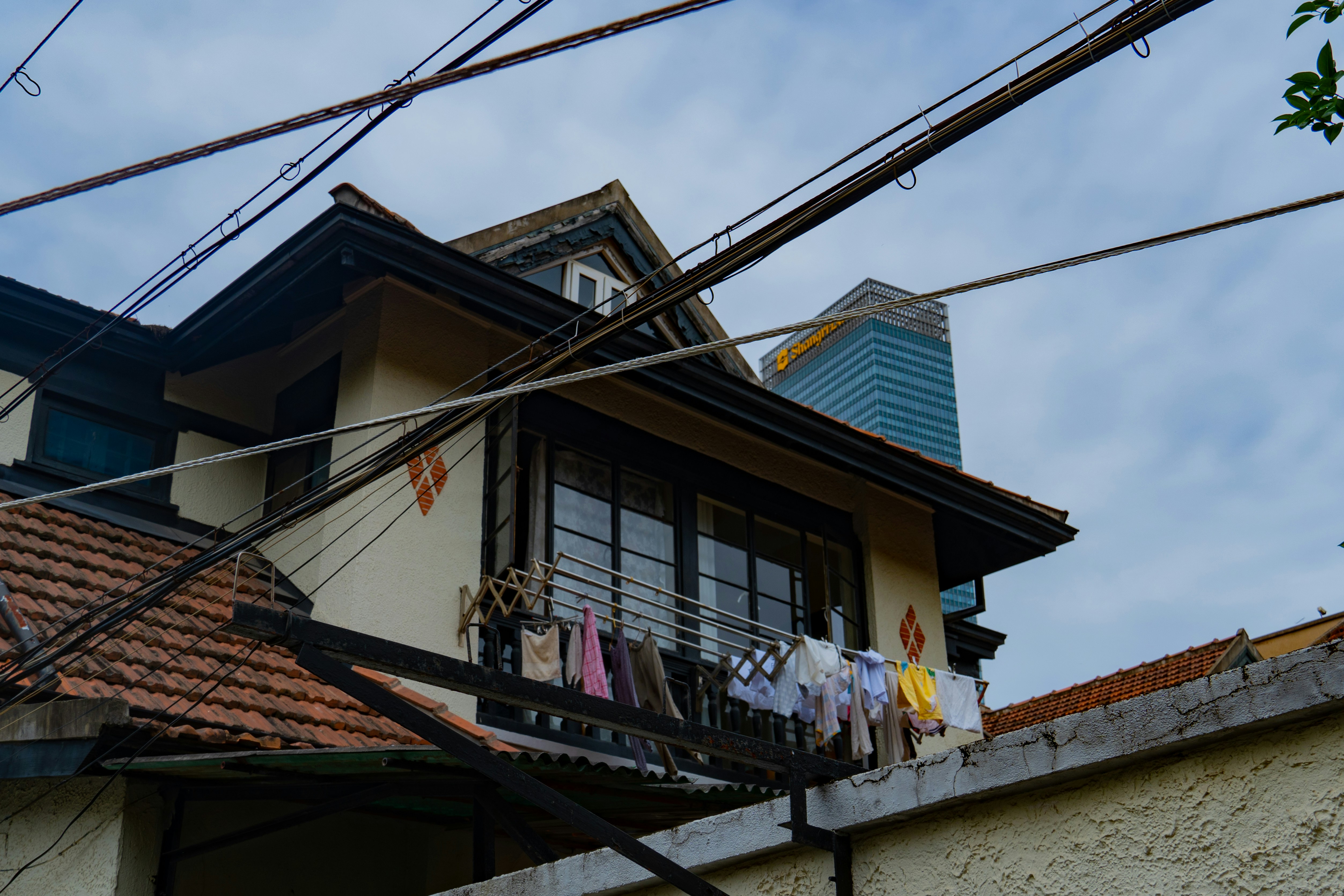 Clothes hanging on a line outside a house with tiled roof and overhead wires against a cloudy sky.