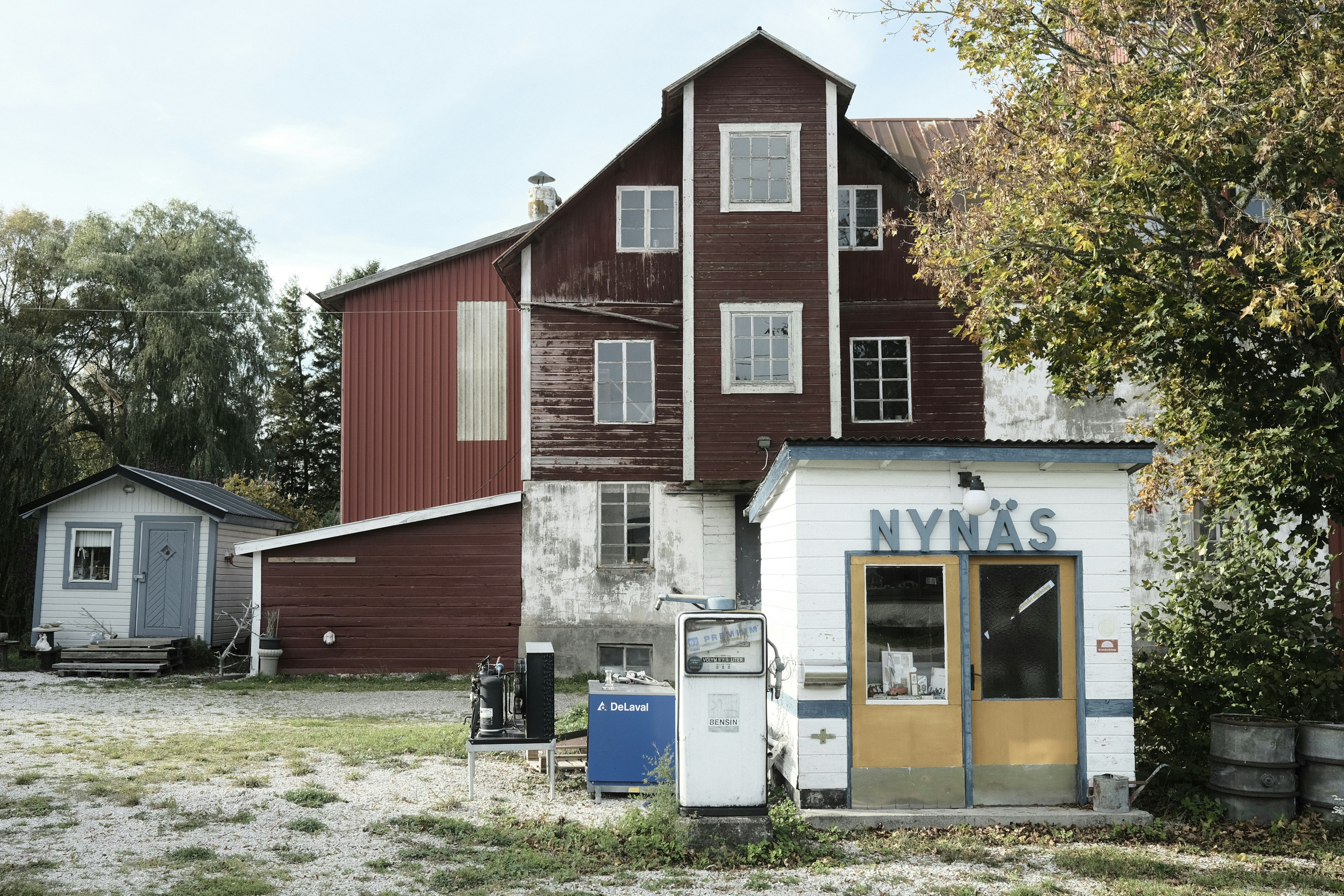 In the front is an old gas pump at gas station with a sign saying 'Nynäs' on it. Behind that, a large red building against a pale blue sky.