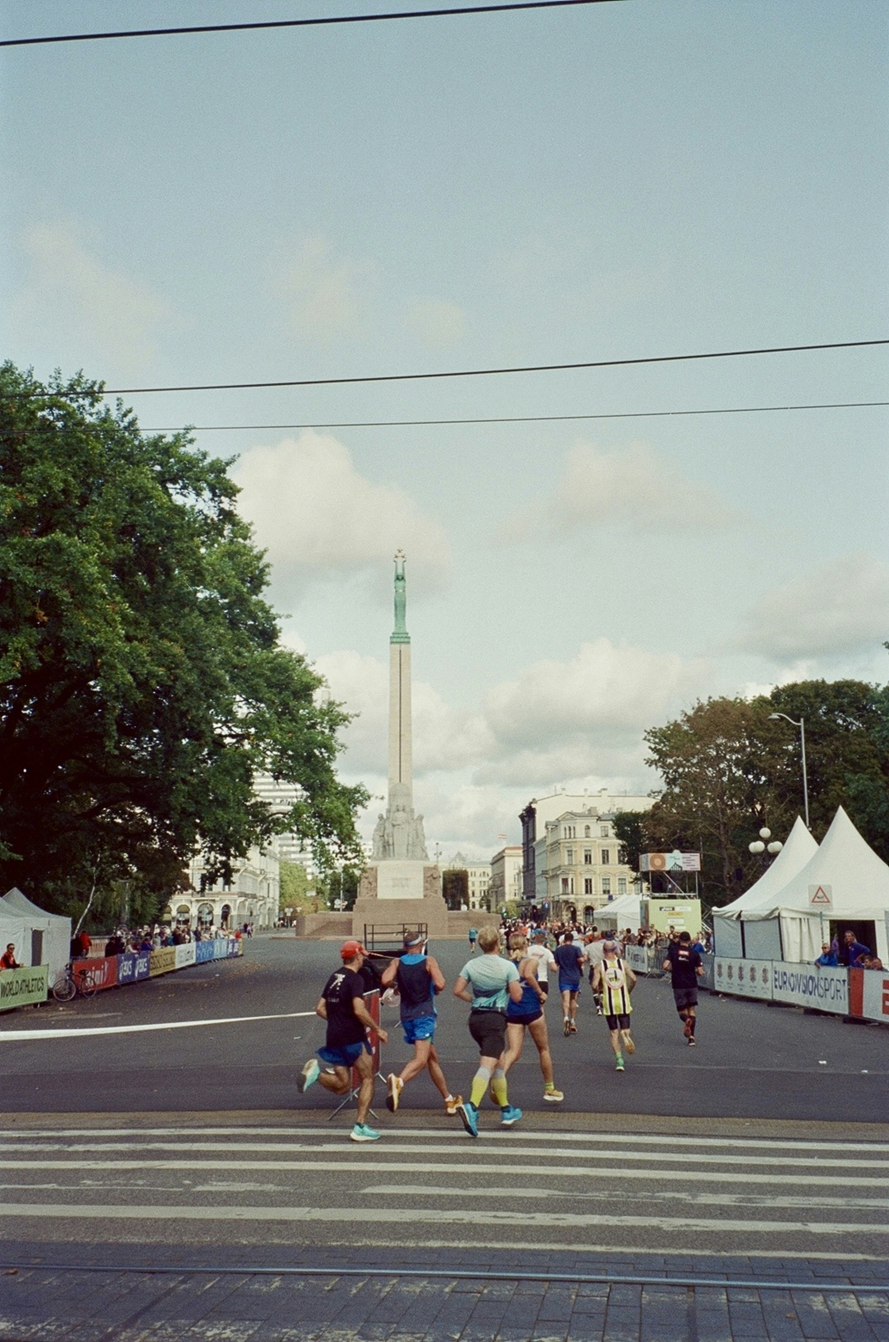 Runners sprint across a city boulevard toward a tall obelisk monument, with tents and spectators lining the street.