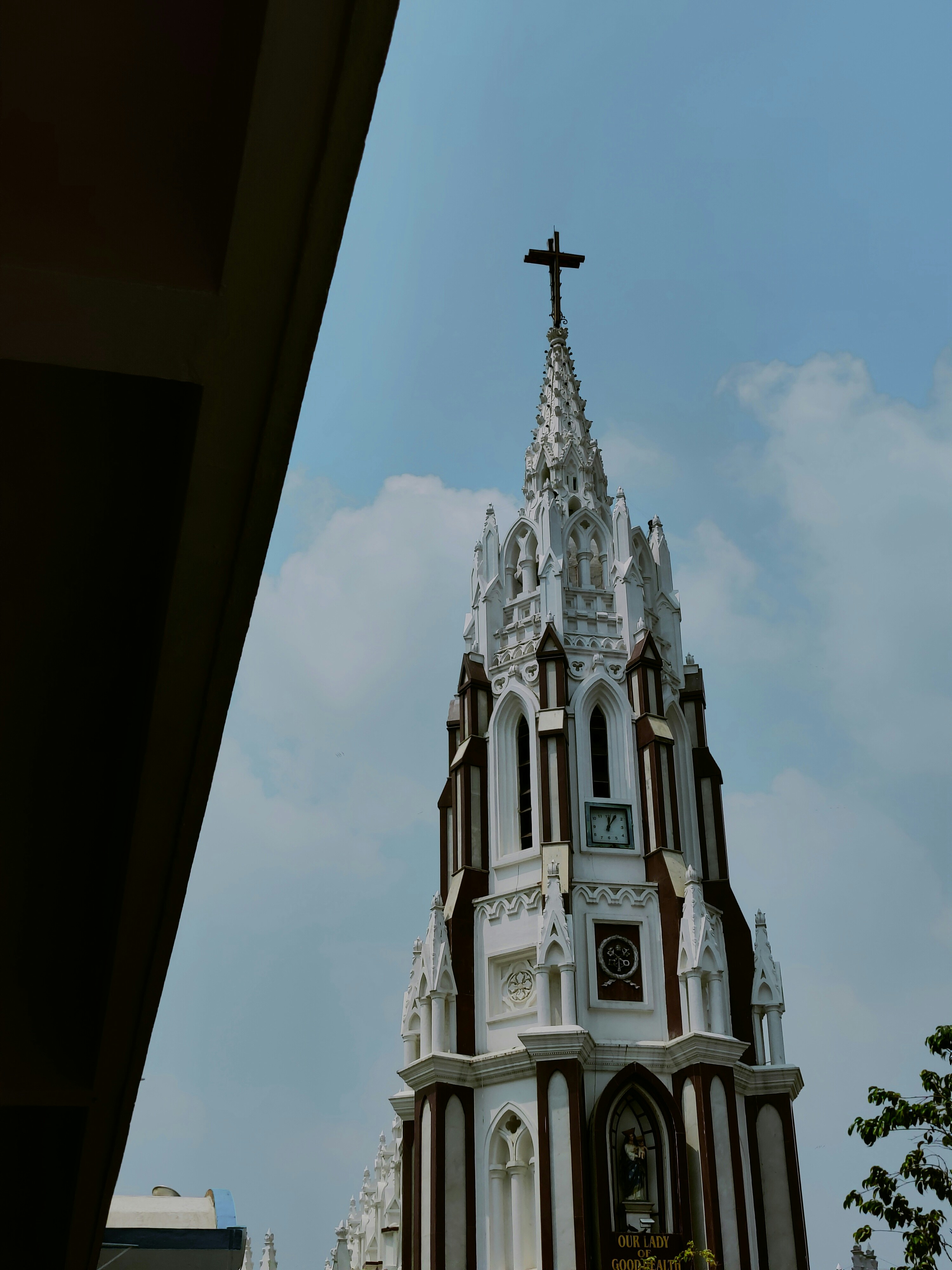 Daylight photograph of a Gothic church spire rising against a pale blue sky, framed by a dark foreground edge.