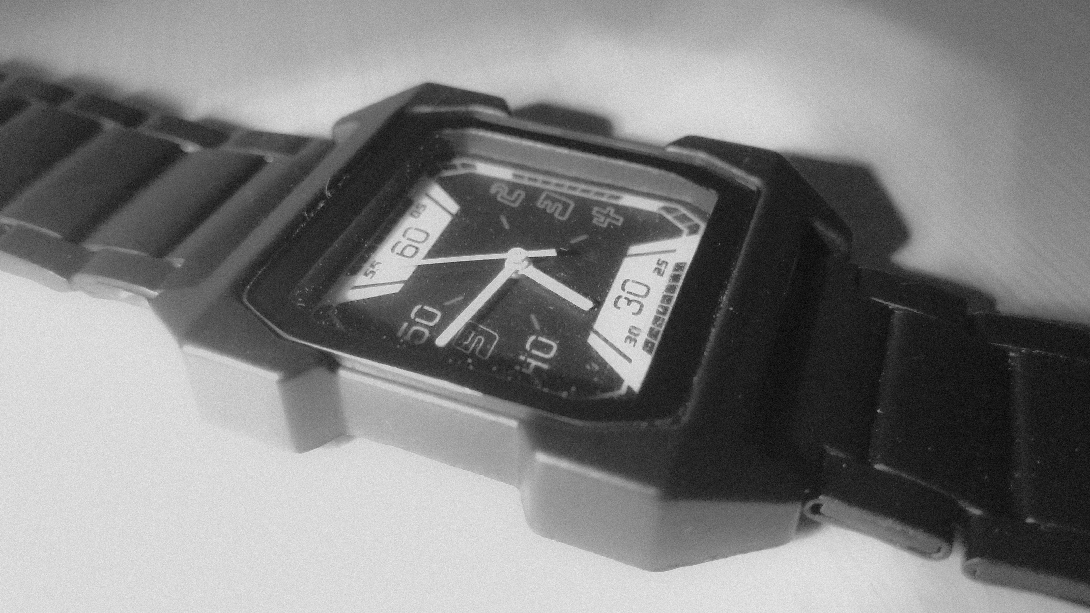 Monochrome close-up of a square-faced wristwatch resting on its metal bracelet on a light surface. The composition emphasizes the dial and angular case with soft shadows.