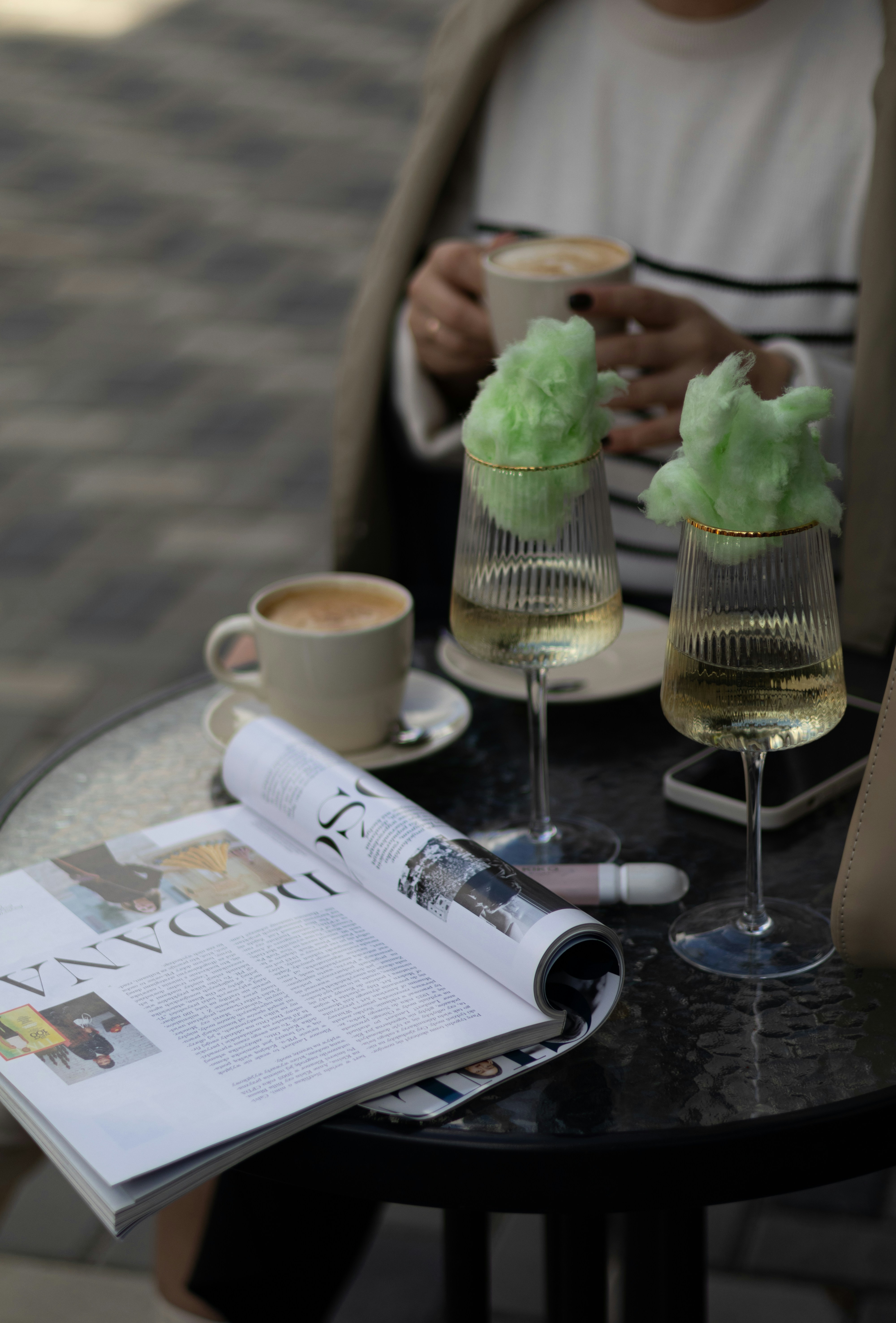 Photograph shows two stemmed wine glasses with pale wine and bright green cotton garnish on a patterned table, an open magazine, and a cup of coffee, with a blurred person in striped top in the background.