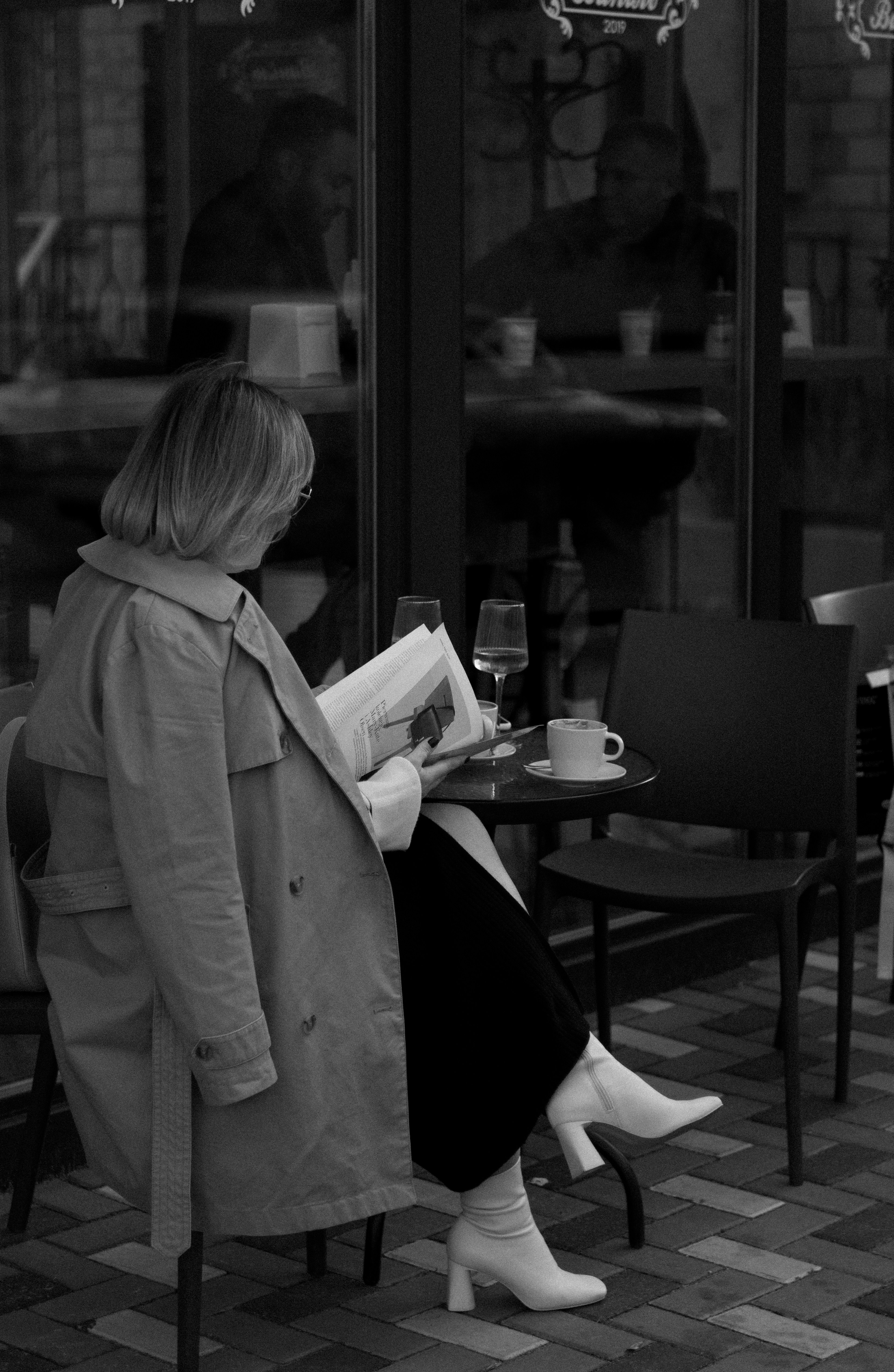 Black-and-white photograph of a woman in a trench coat reading a book at a small café table, with a cup and a wine glass nearby.